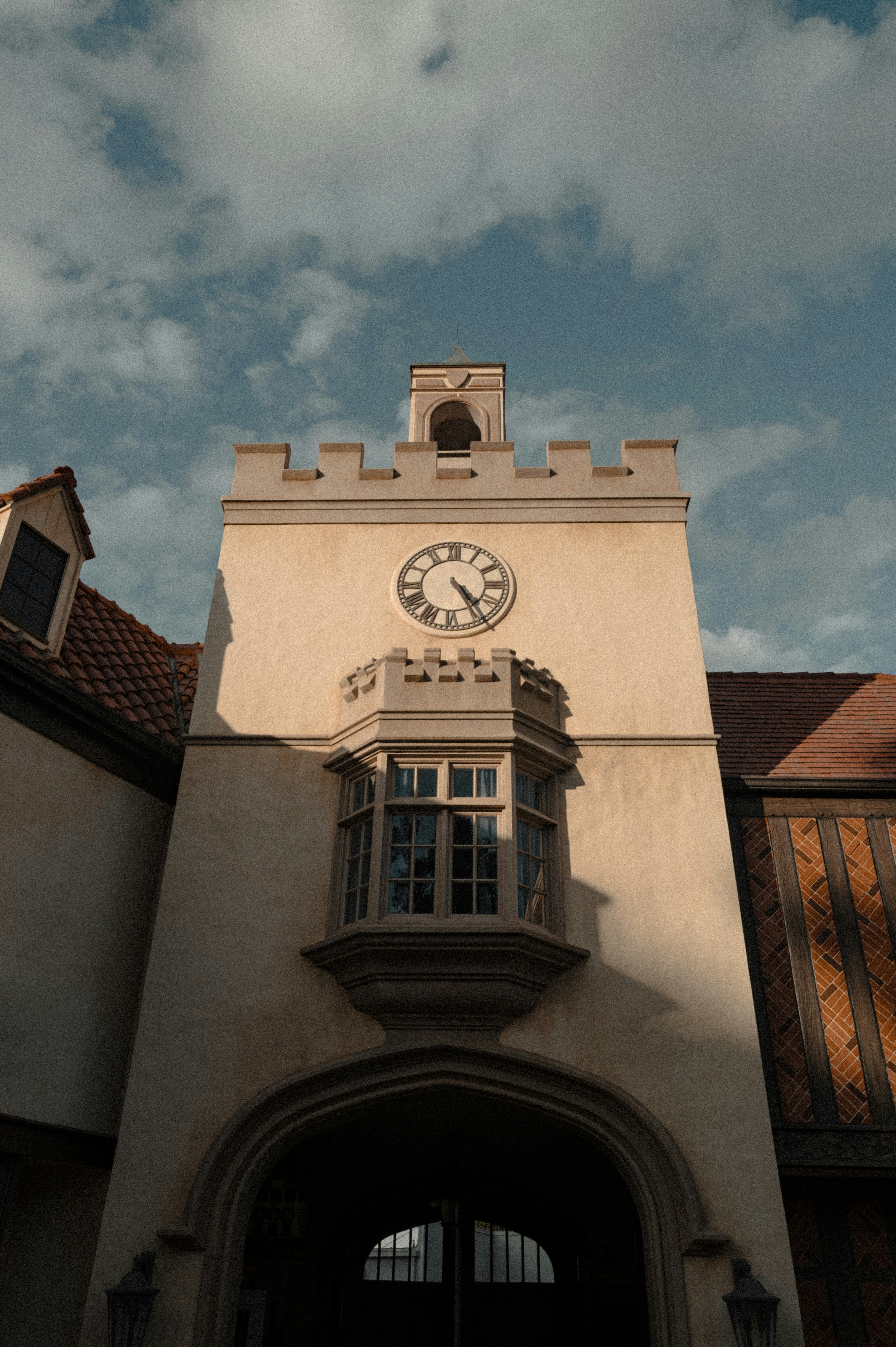 Medieval-style gatehouse with a centered clock tower, bay window, and crenellated parapets rising over a rounded arch against a bright blue sky.