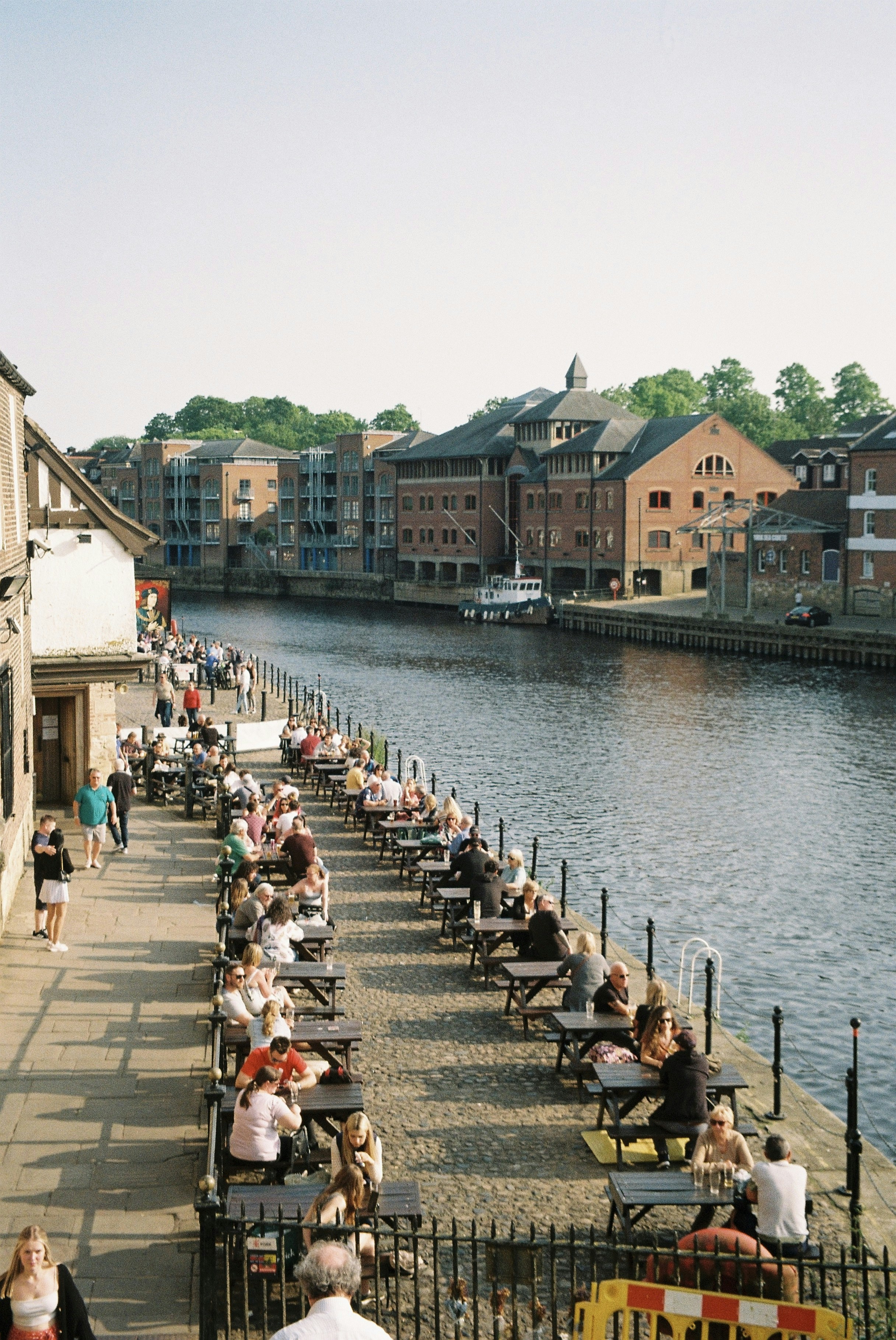 People enjoying a sunny day at outdoor tables along a riverbank, with brick buildings and lush trees in the background.