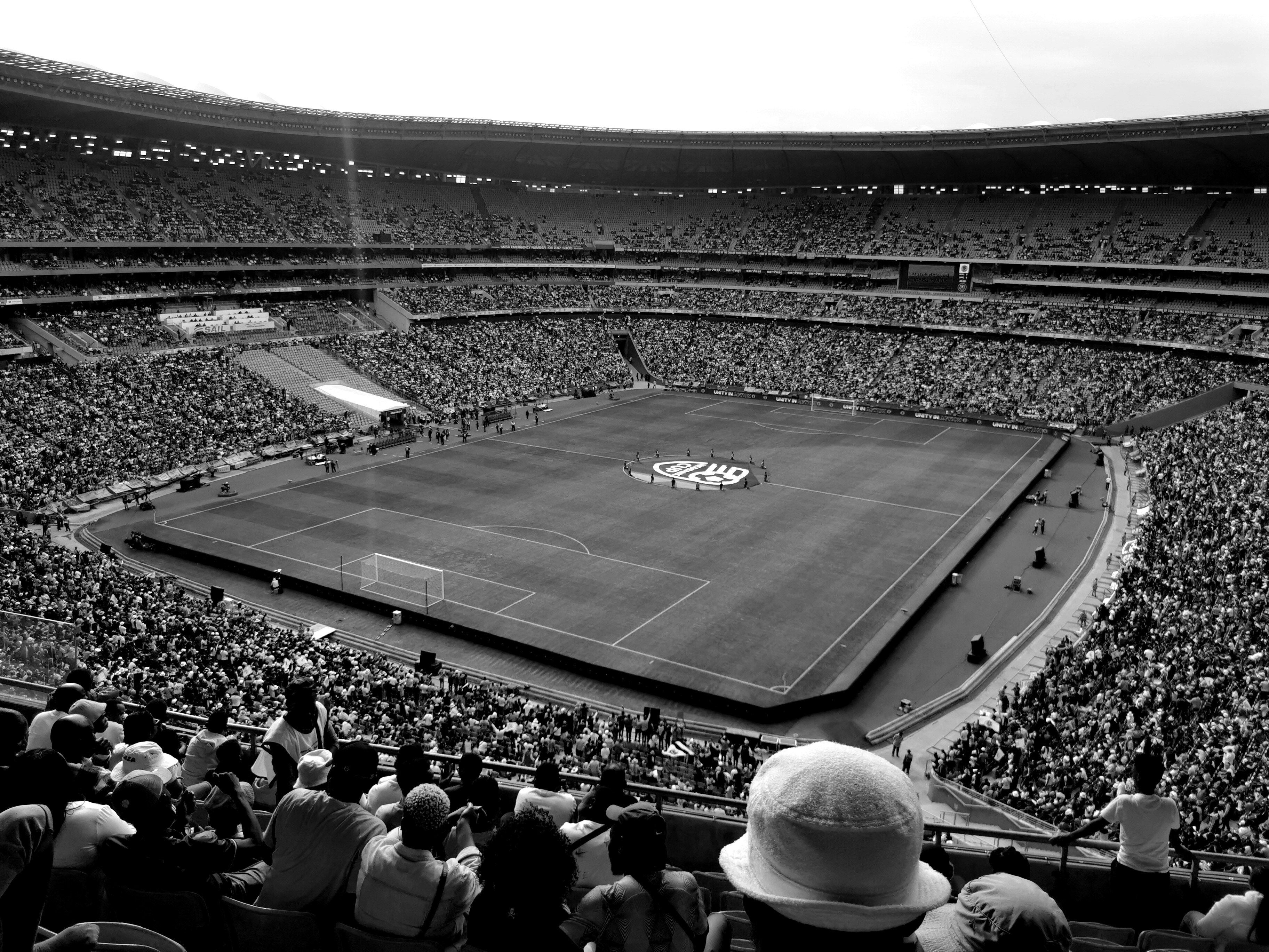 Wide-angle view of a packed stadium with spectators focused on a central field under bright daylight.