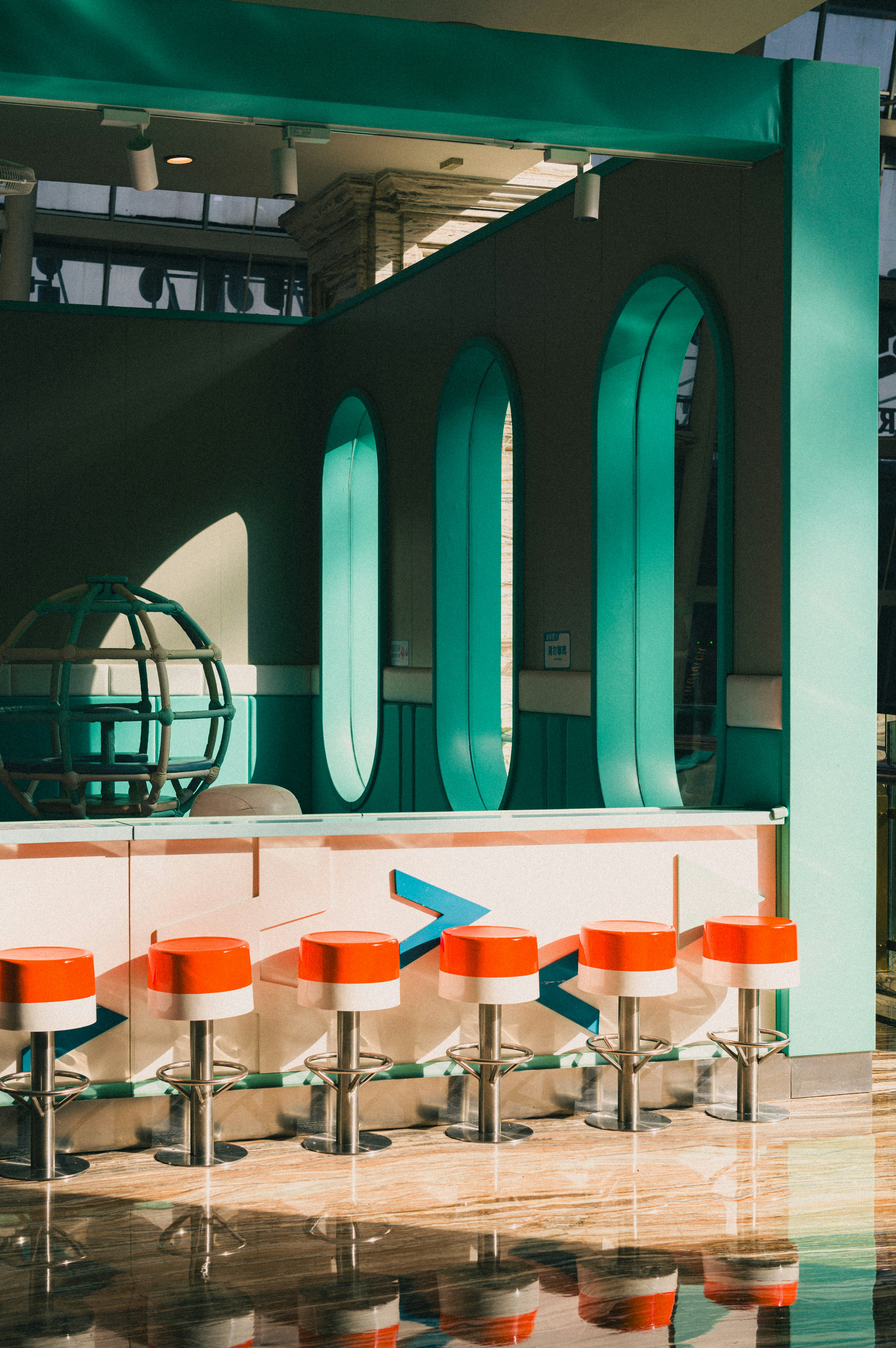 Photograph of a retro cafe interior with turquoise arches and a row of orange-topped stools. The glossy floor reflects the scene, highlighting color and architectural rhythm.
