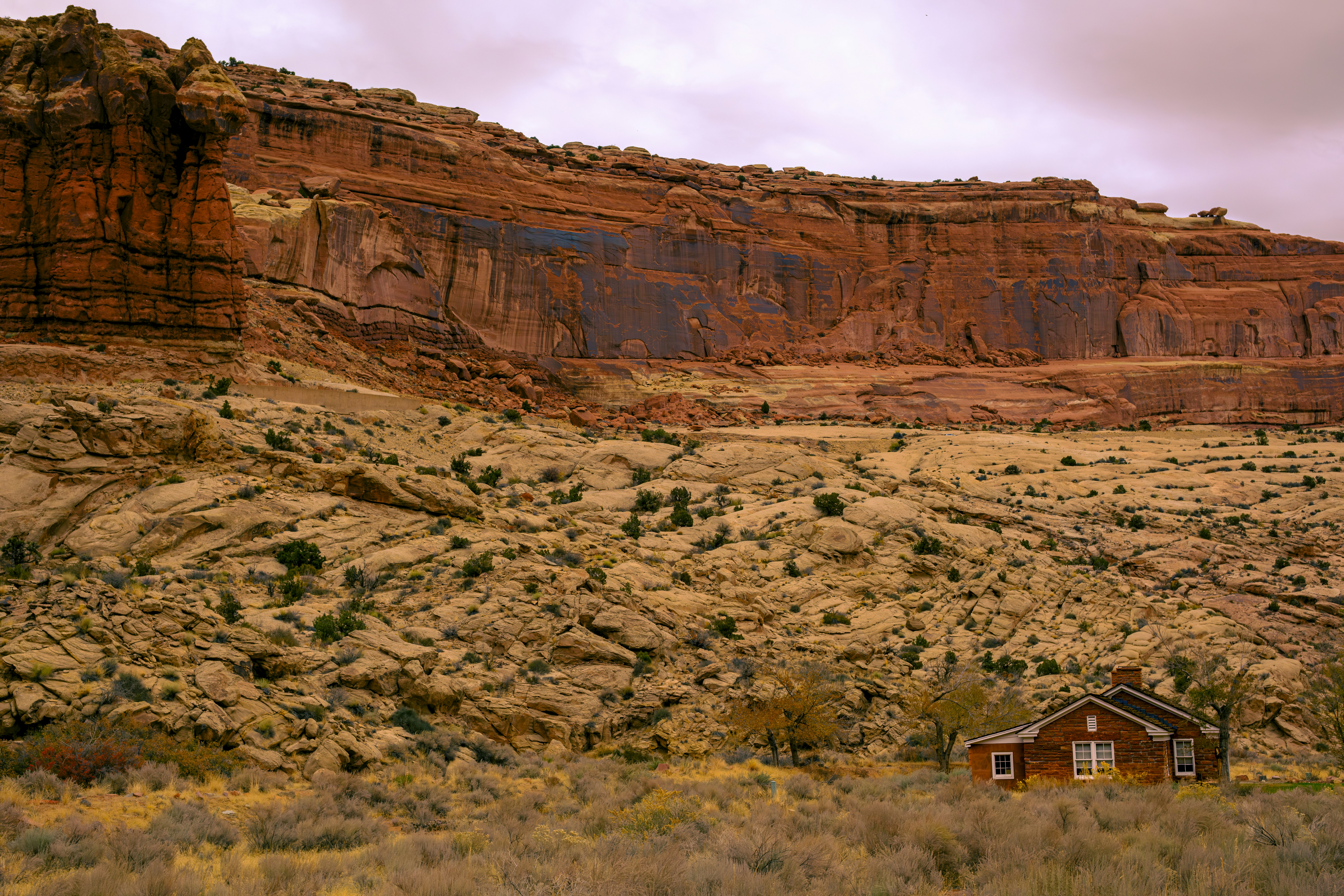 Small cabin nestled in a vast desert landscape under towering red cliffs.