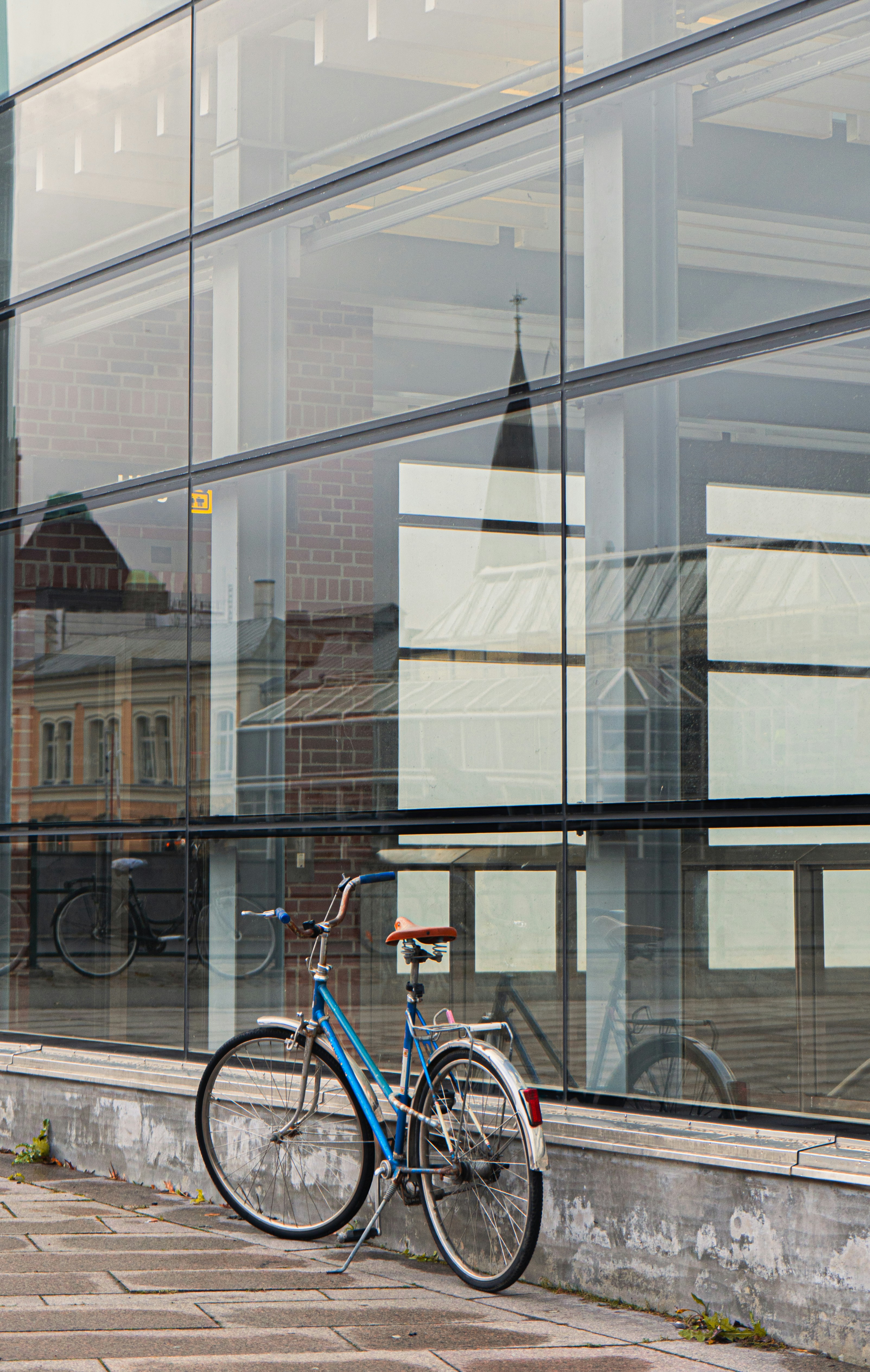 A blue bicycle rests against a modern glass facade, reflecting the surrounding architecture and capturing the essence of urban life.