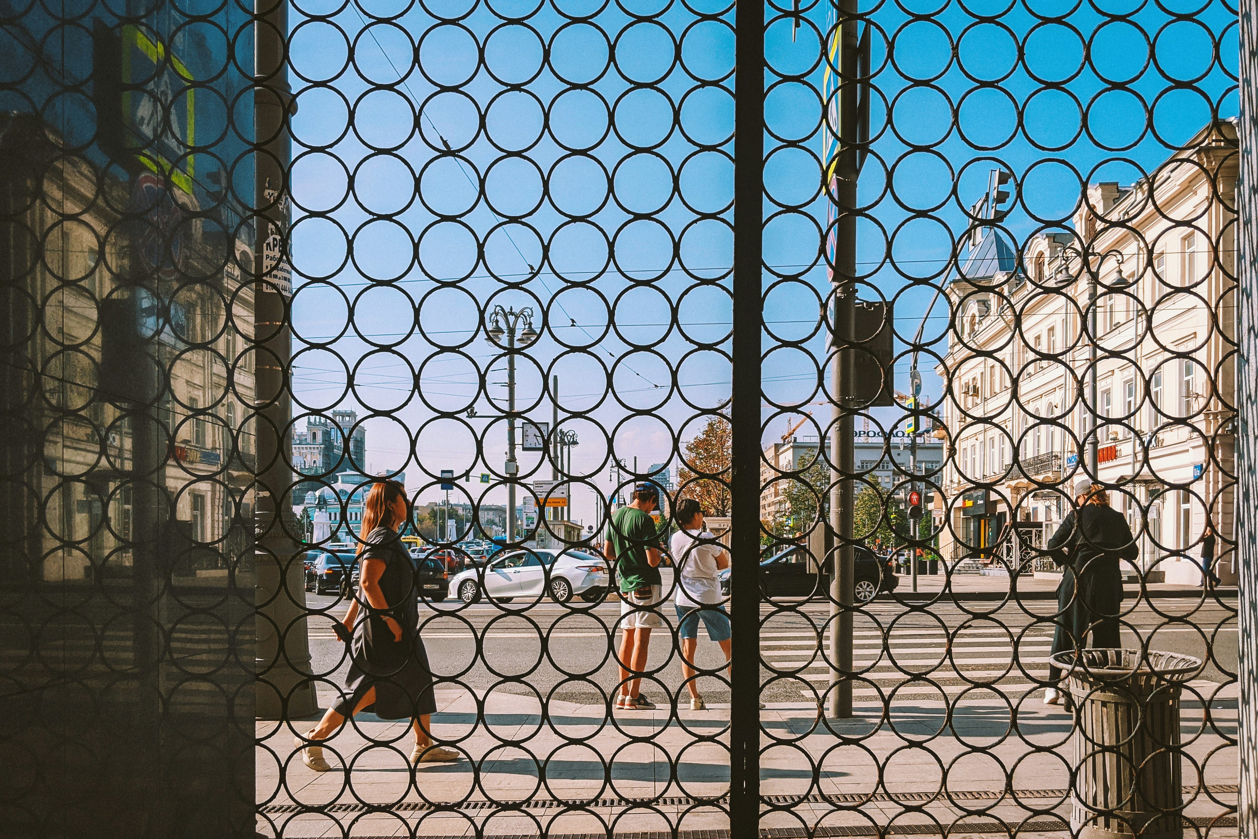 Framed in Circles: City Pedestrians Walk a Sunlit Plaza Behind a Grille