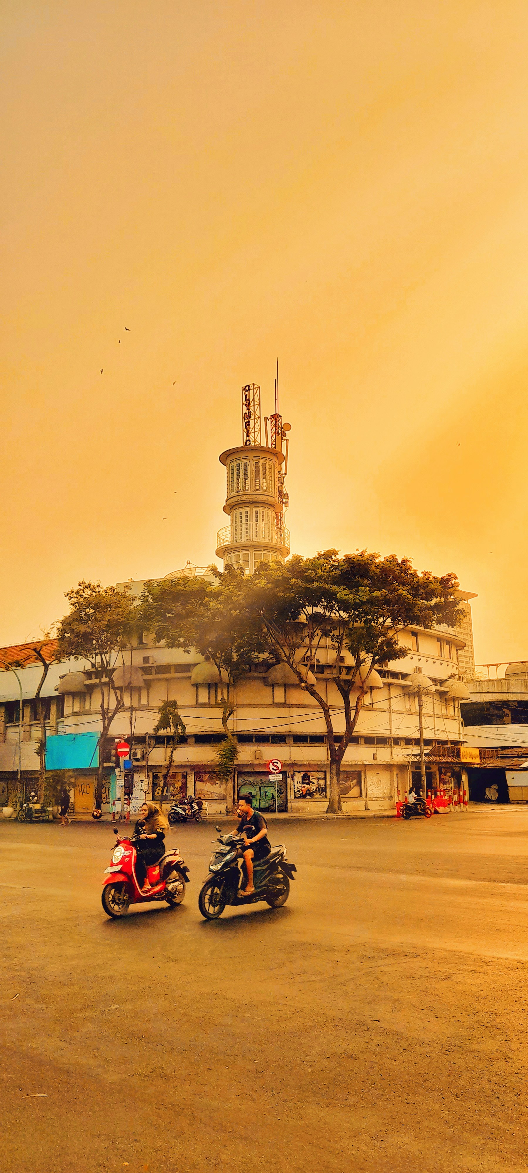 Street scene at dusk featuring a cylindrical tower with antennas atop a low-rise building, framed by trees and scooters.