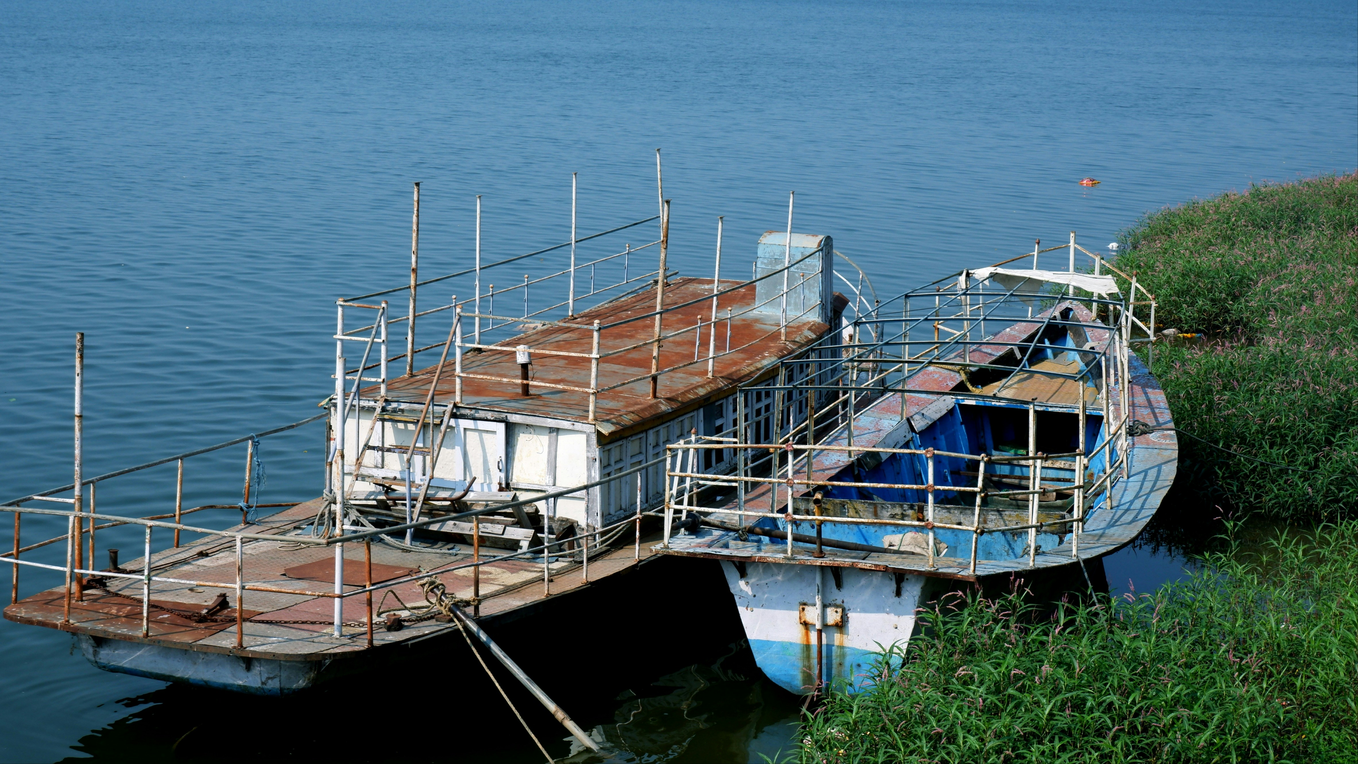 Photograph of two weathered boats moored along a calm blue waterway, their decks rusted and railings weather-beaten.
