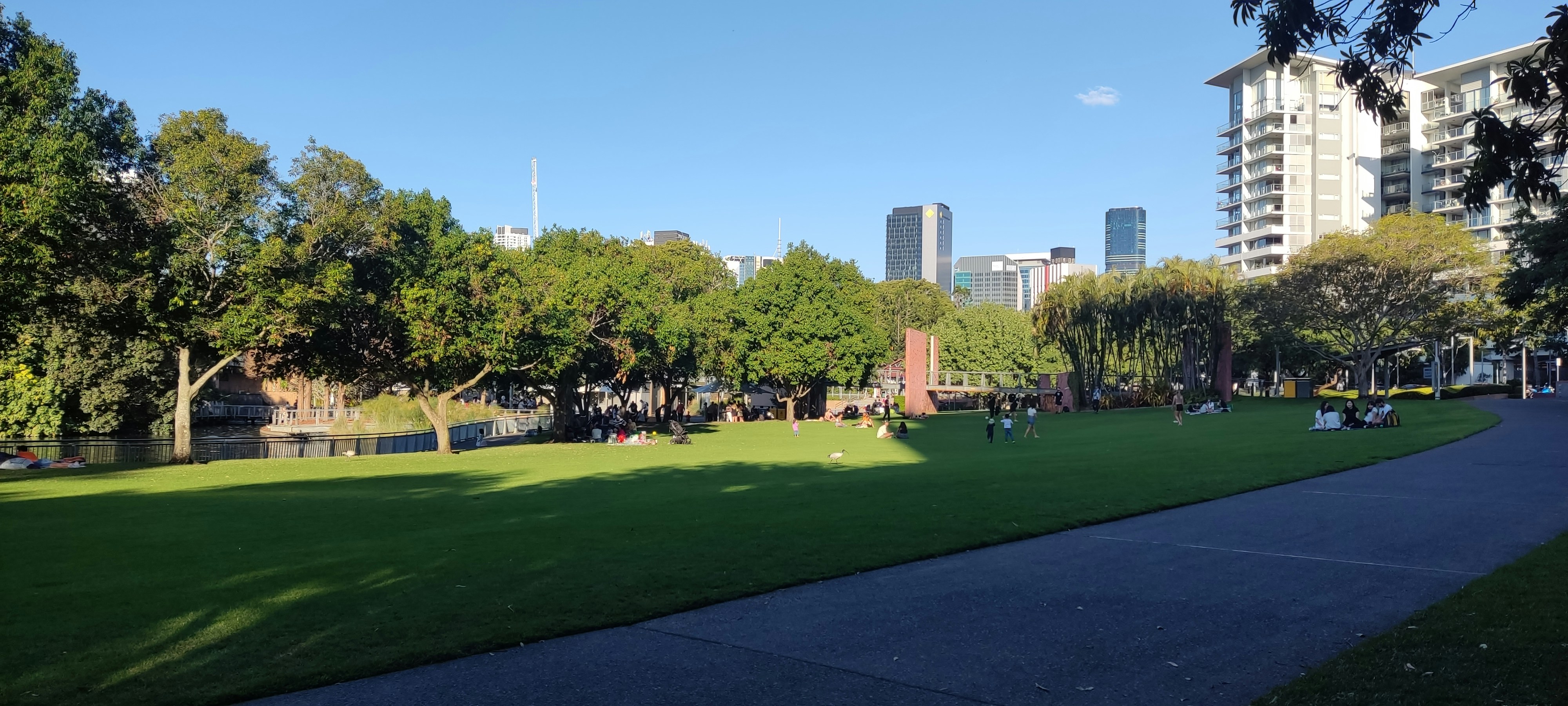 Photograph of a sunlit city park featuring a brick obelisk along a paved path and groups of people on the grass, with distant skyline buildings.