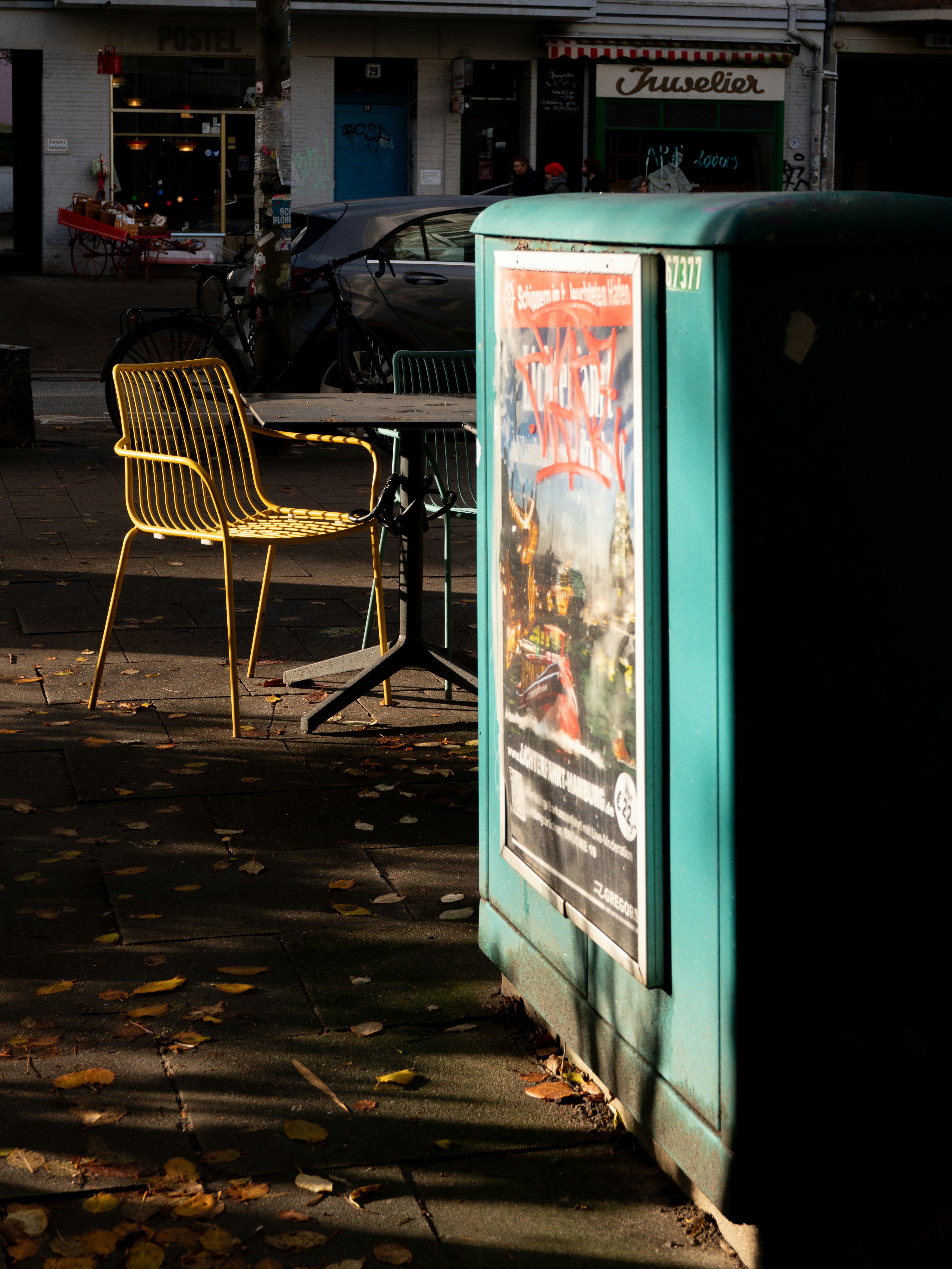 Sunlit Geometry: A Yellow Chair Befriends a Teal Newsstand on a Quiet Street