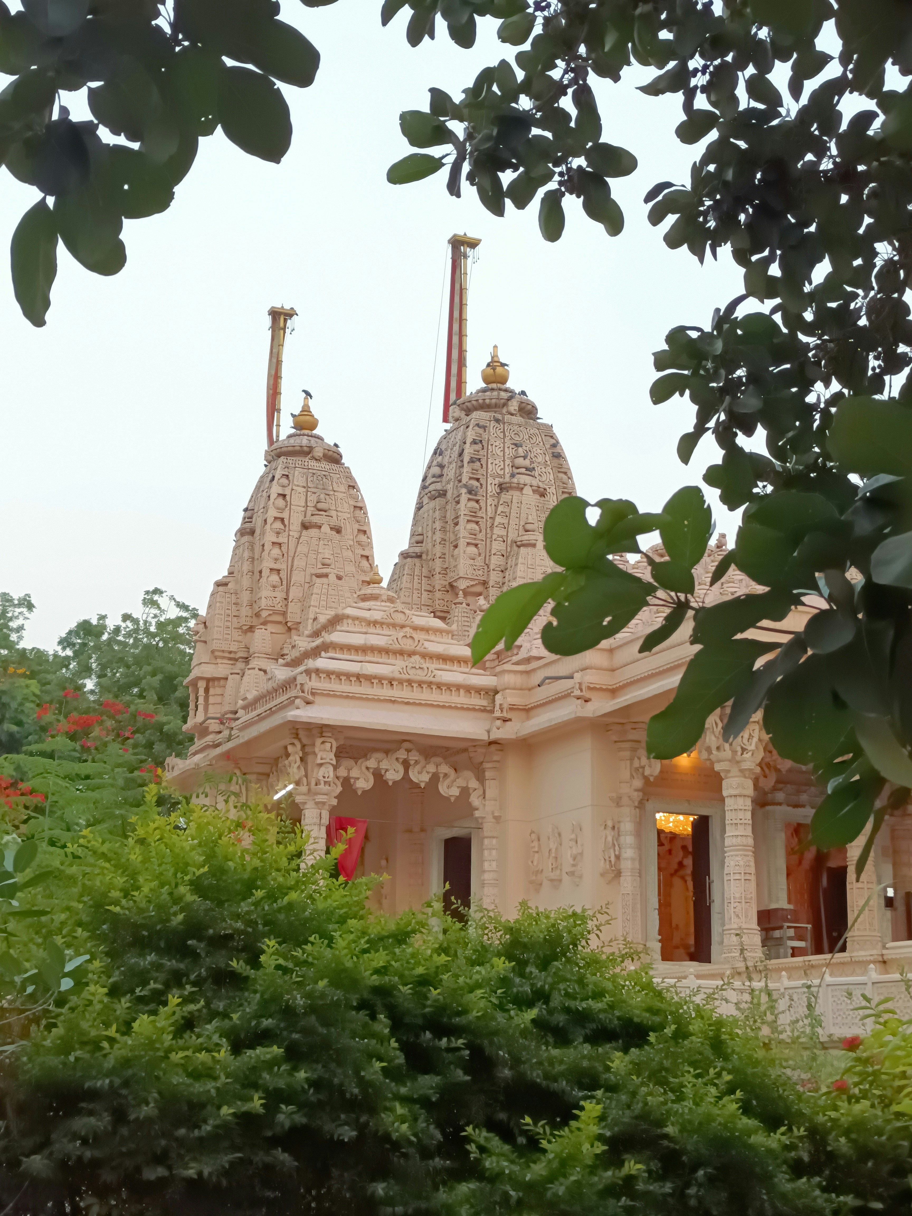 Ornate stone temple spires rise from lush greenery, framed by overhanging leaves. The scene highlights intricate carvings and a serene garden setting.