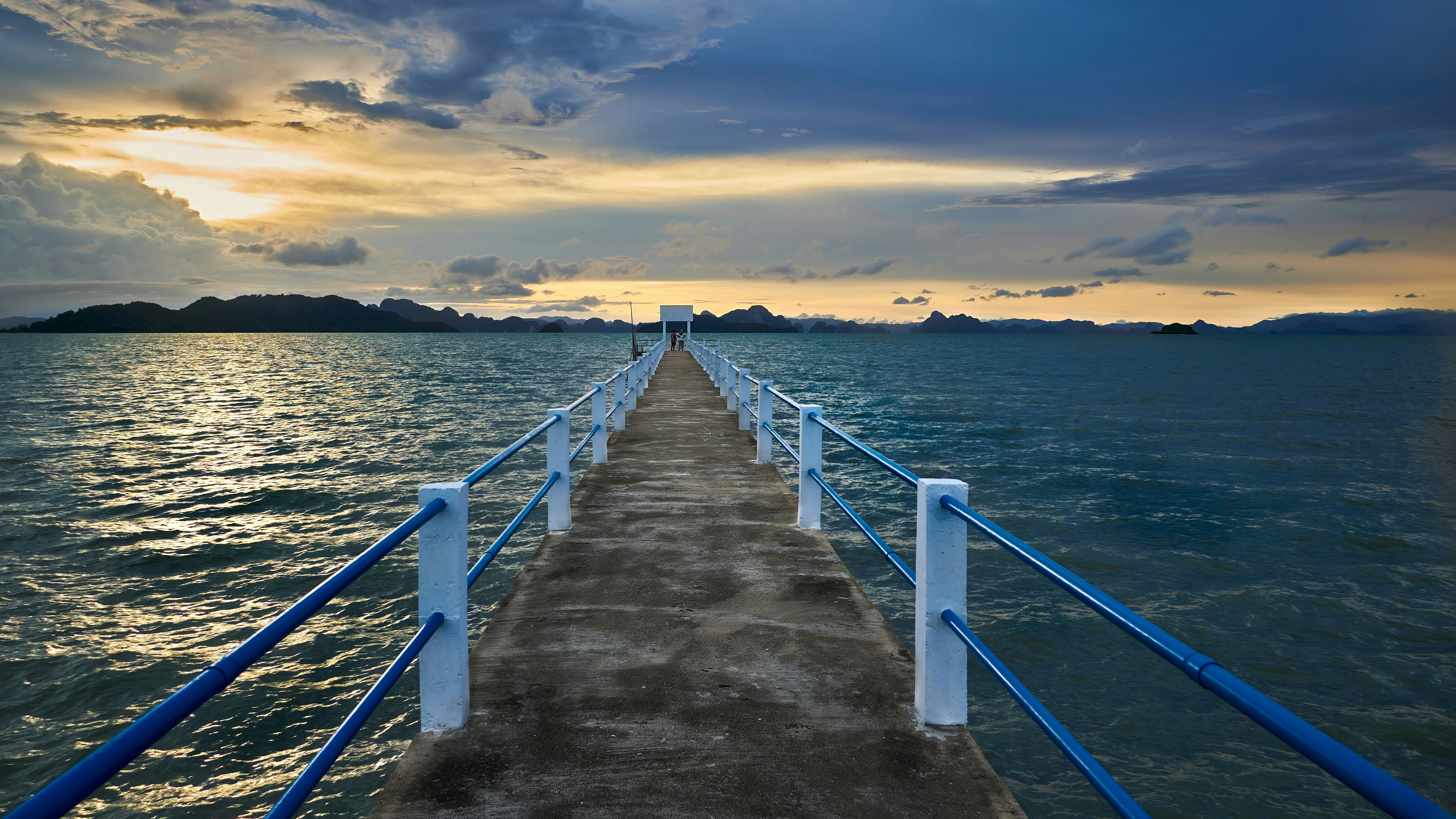 Concrete pier stretching into the ocean beneath a dramatic cloudy sky at sunset.