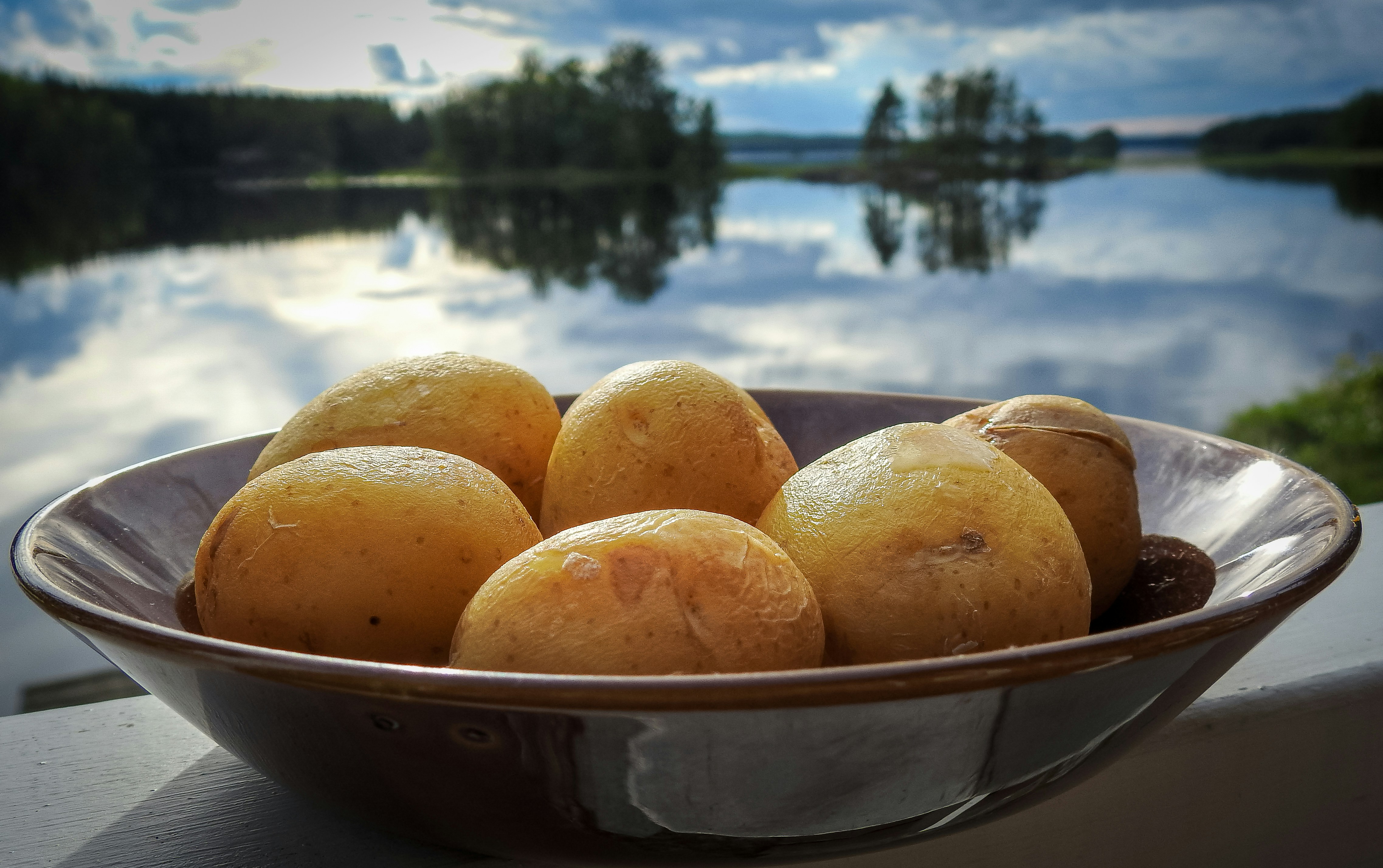 Photograph of golden potatoes in a rustic ceramic bowl resting on a lakeside table, with a mirror-like lake and trees reflected in the background.