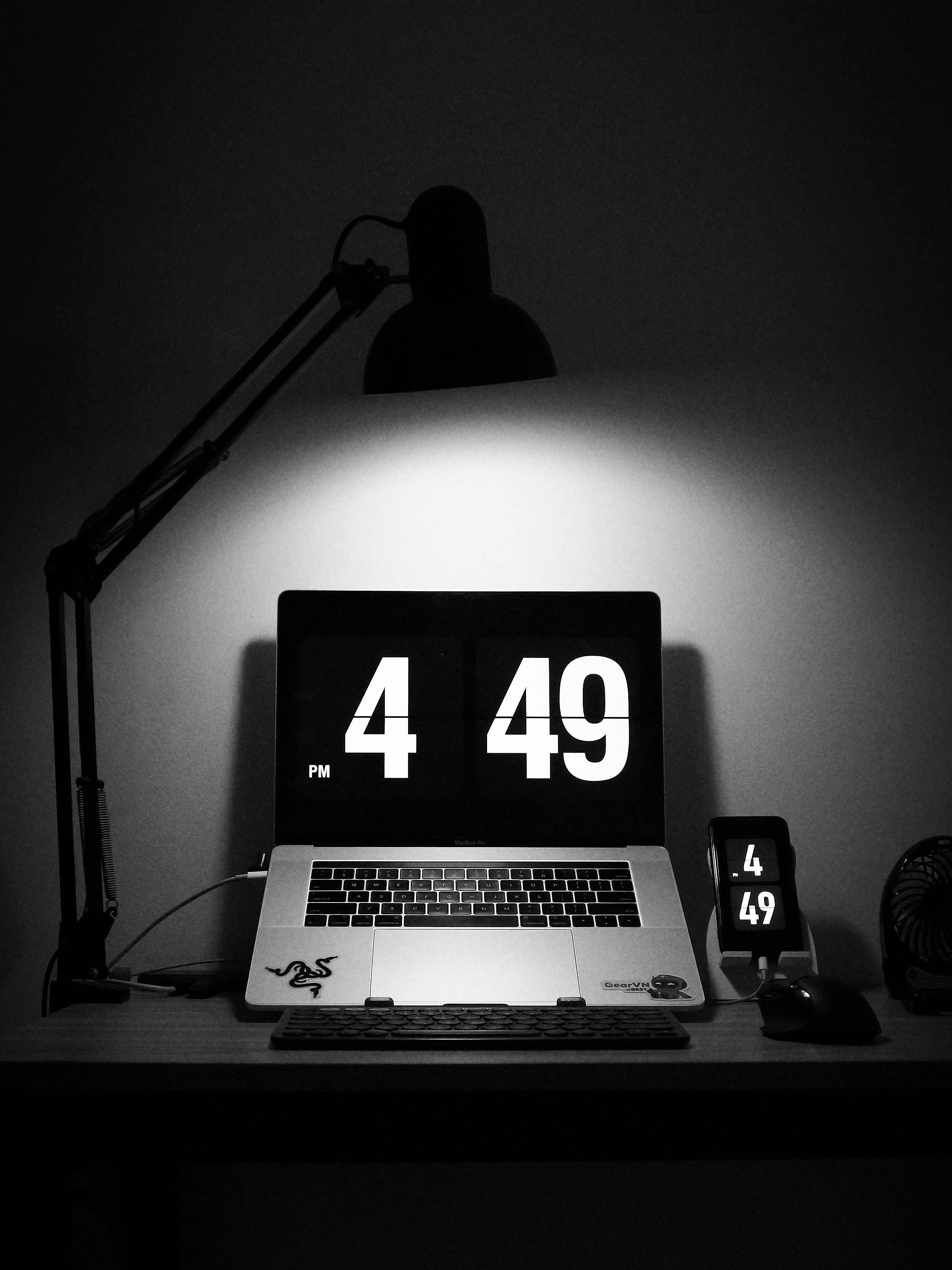 A minimalist workspace featuring a laptop displaying the time '4:49 PM' alongside a smartphone showing the same time, all illuminated by a desk lamp.