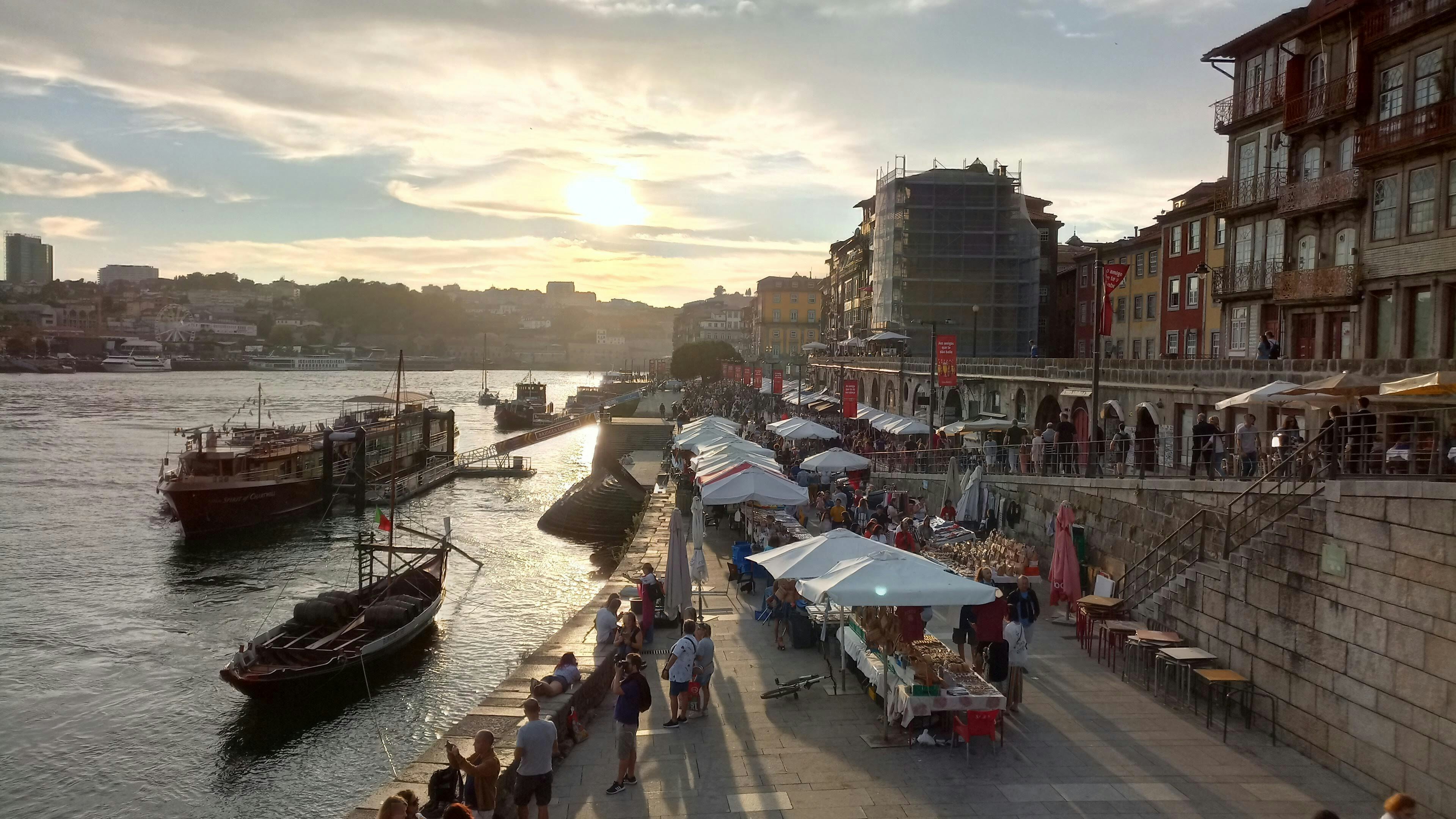 People walking along a riverside promenade with market stalls and boats under a setting sun.