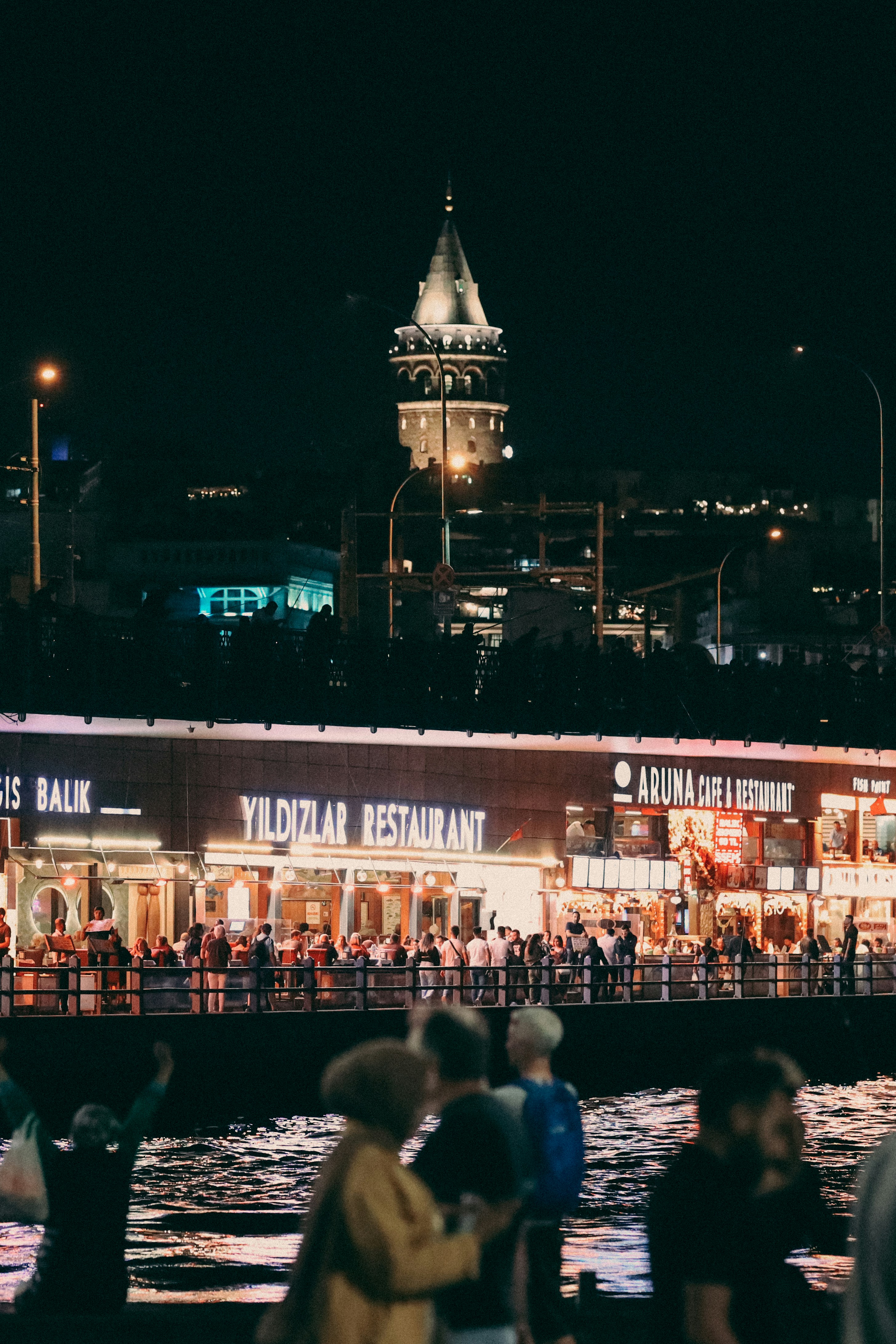 Illuminated waterfront restaurants bustling with diners under the night sky, featuring a historic tower in the background.
