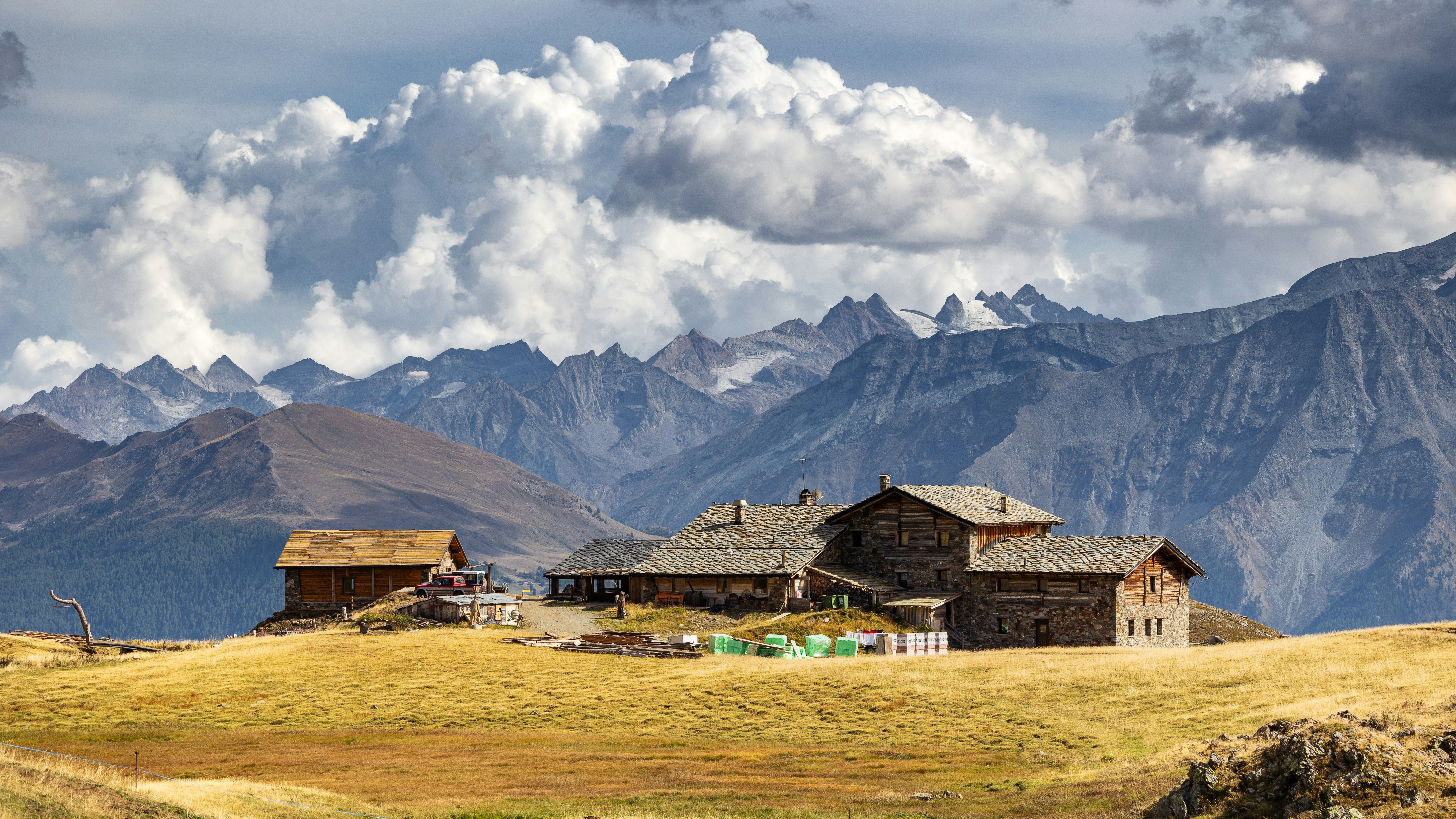 Rustic houses in a golden field with towering mountains under dramatic clouds.