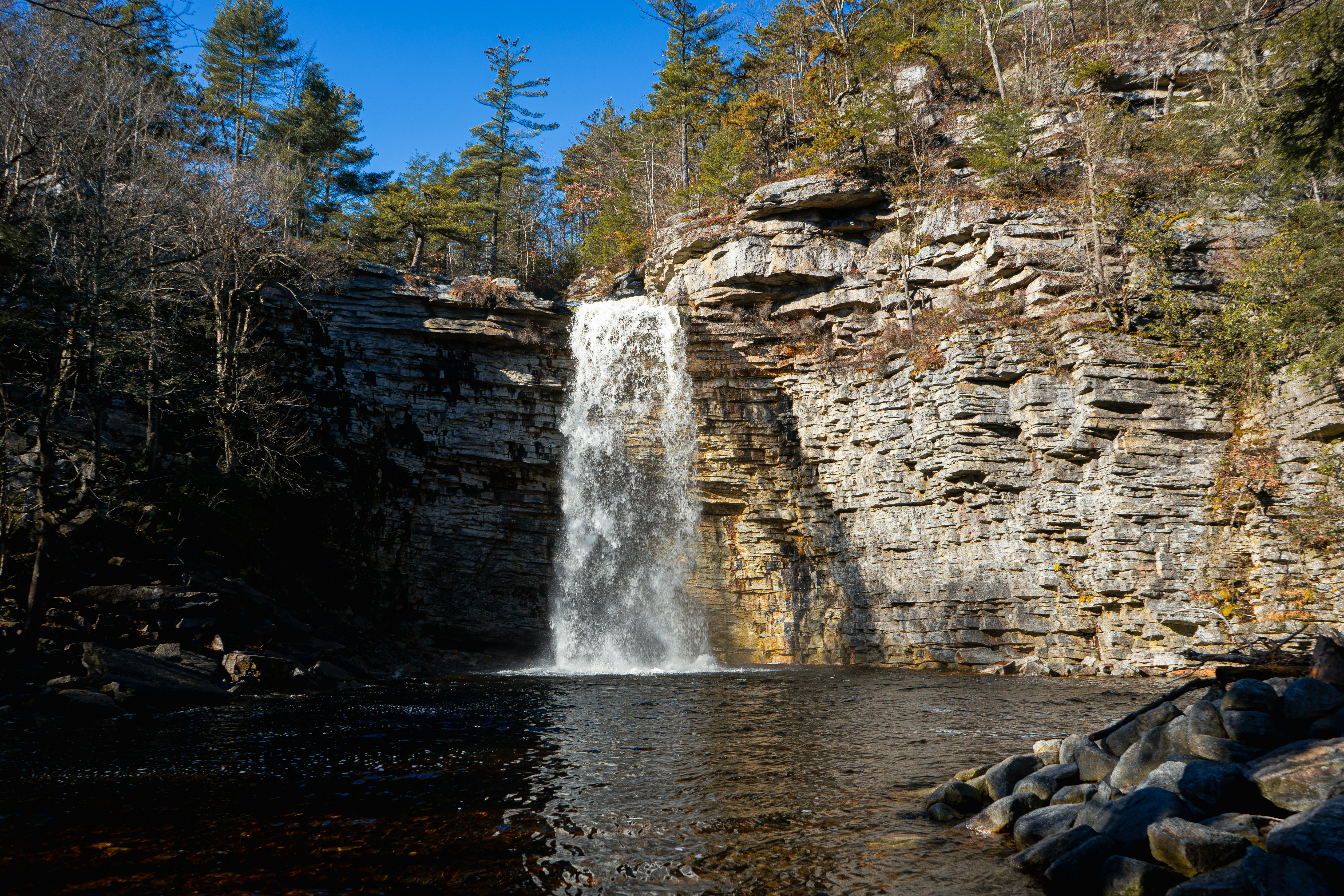 Waterfall cascading down rugged cliffs into a tranquil pool, surrounded by lush forest under a clear blue sky.
