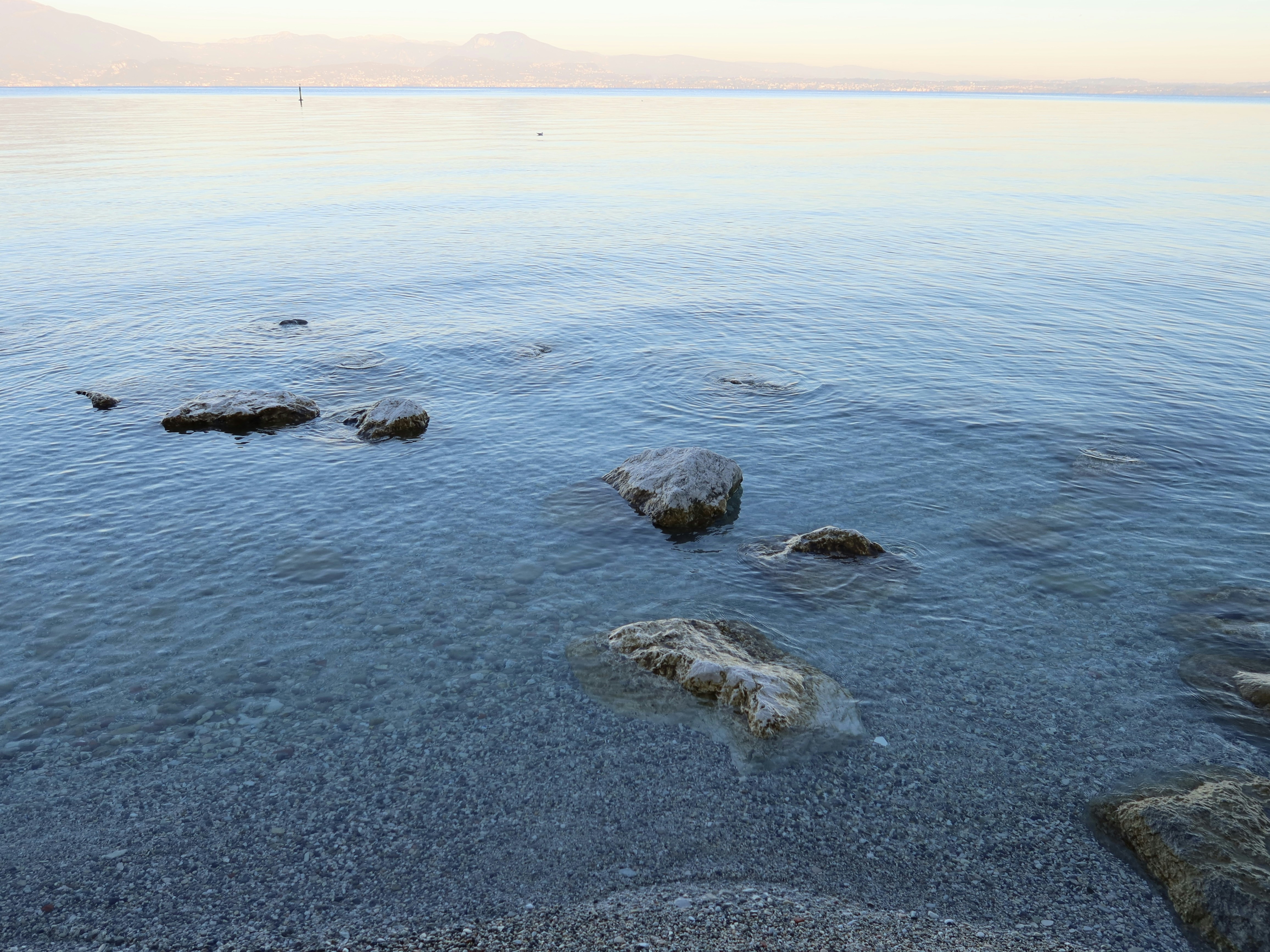 Clear water with rocks submerged near the shore, distant mountains on the horizon under a soft sky.