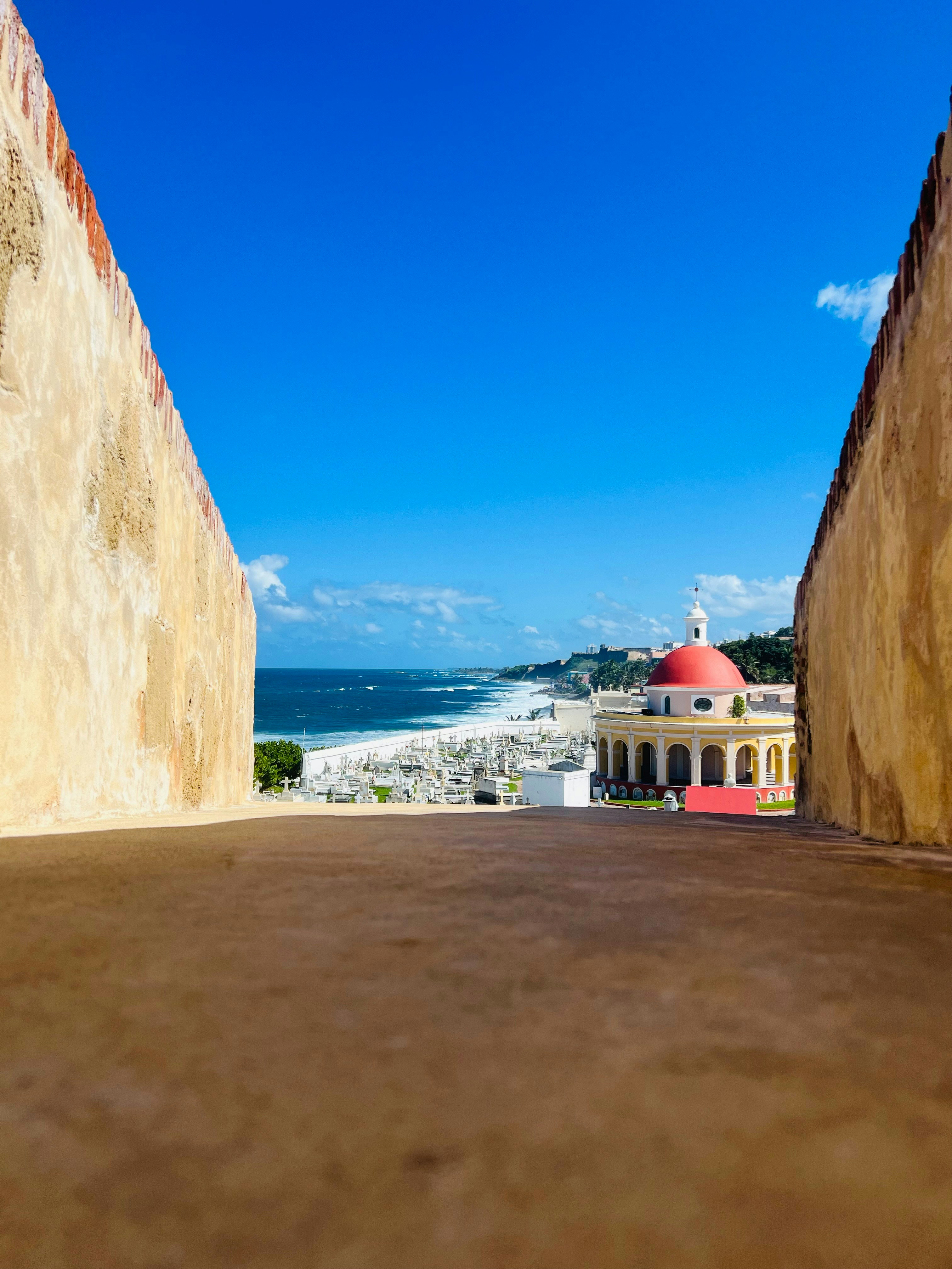 Ocean framed by historic stone walls with a vibrant domed building and cemetery in the distance under a clear blue sky.