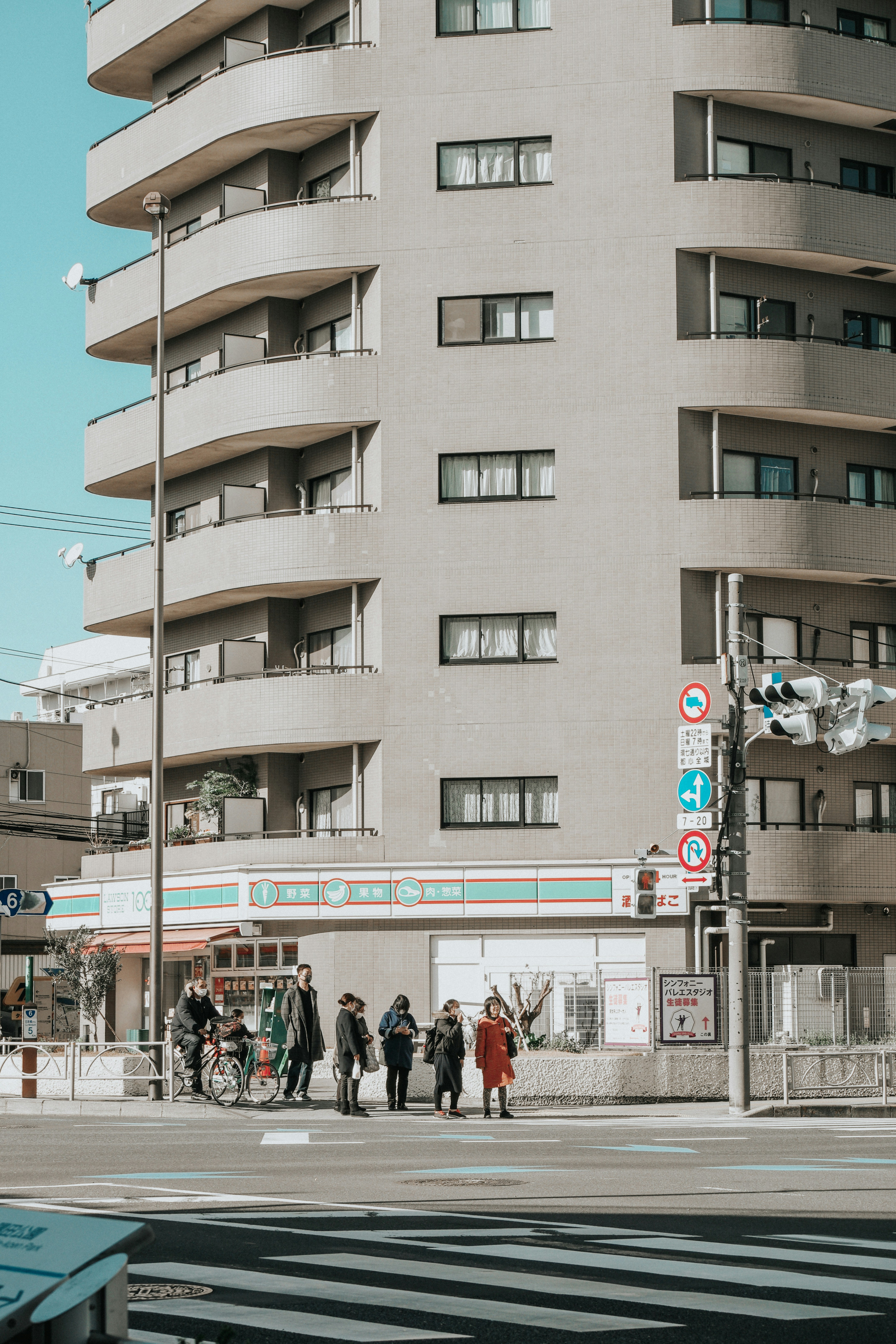Group of pedestrians waiting at a crosswalk in front of a modern building, with a cyclist in the foreground. Traffic signals and signage add to the urban atmosphere.