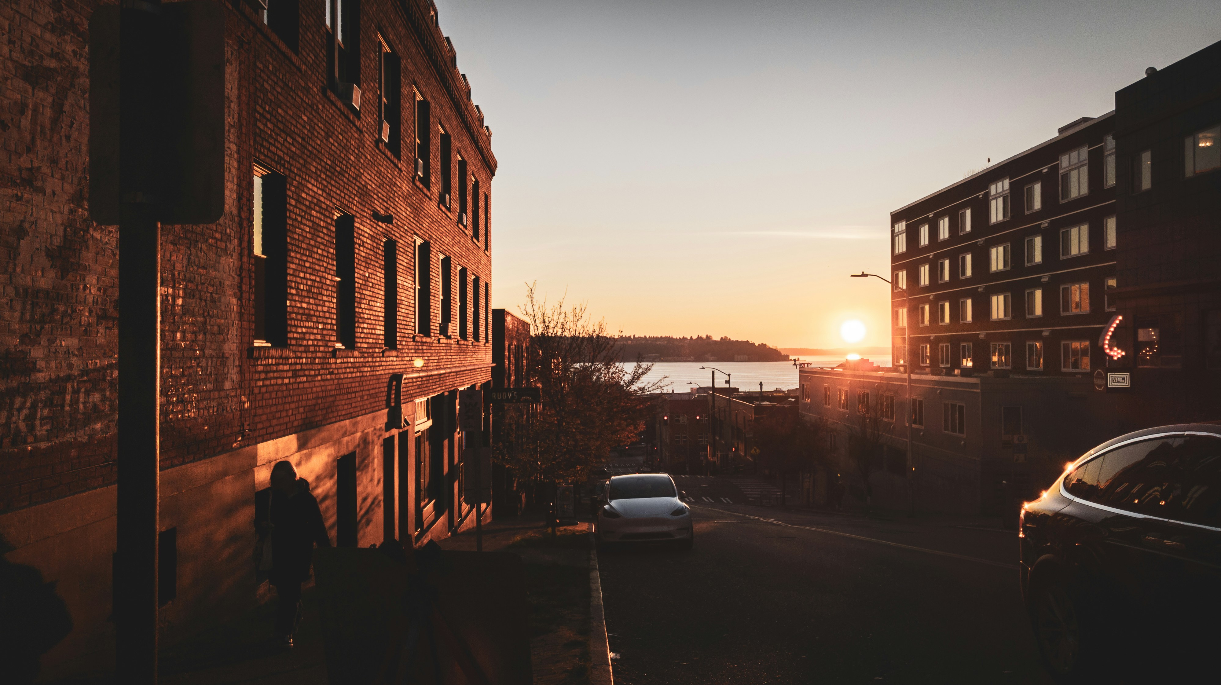 Brick buildings flank a quiet street as the sun sets over a distant water body, bathing the scene in warm orange light.