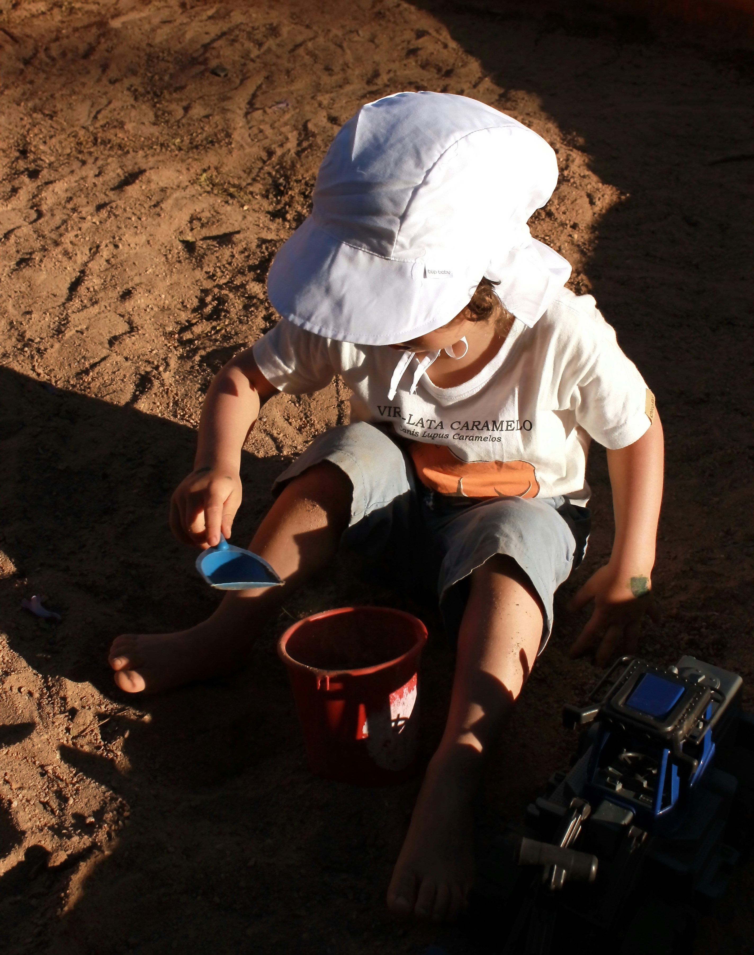 Sunlit Sandbox Play: A toddler in a white hat with a blue scoop and red bucket