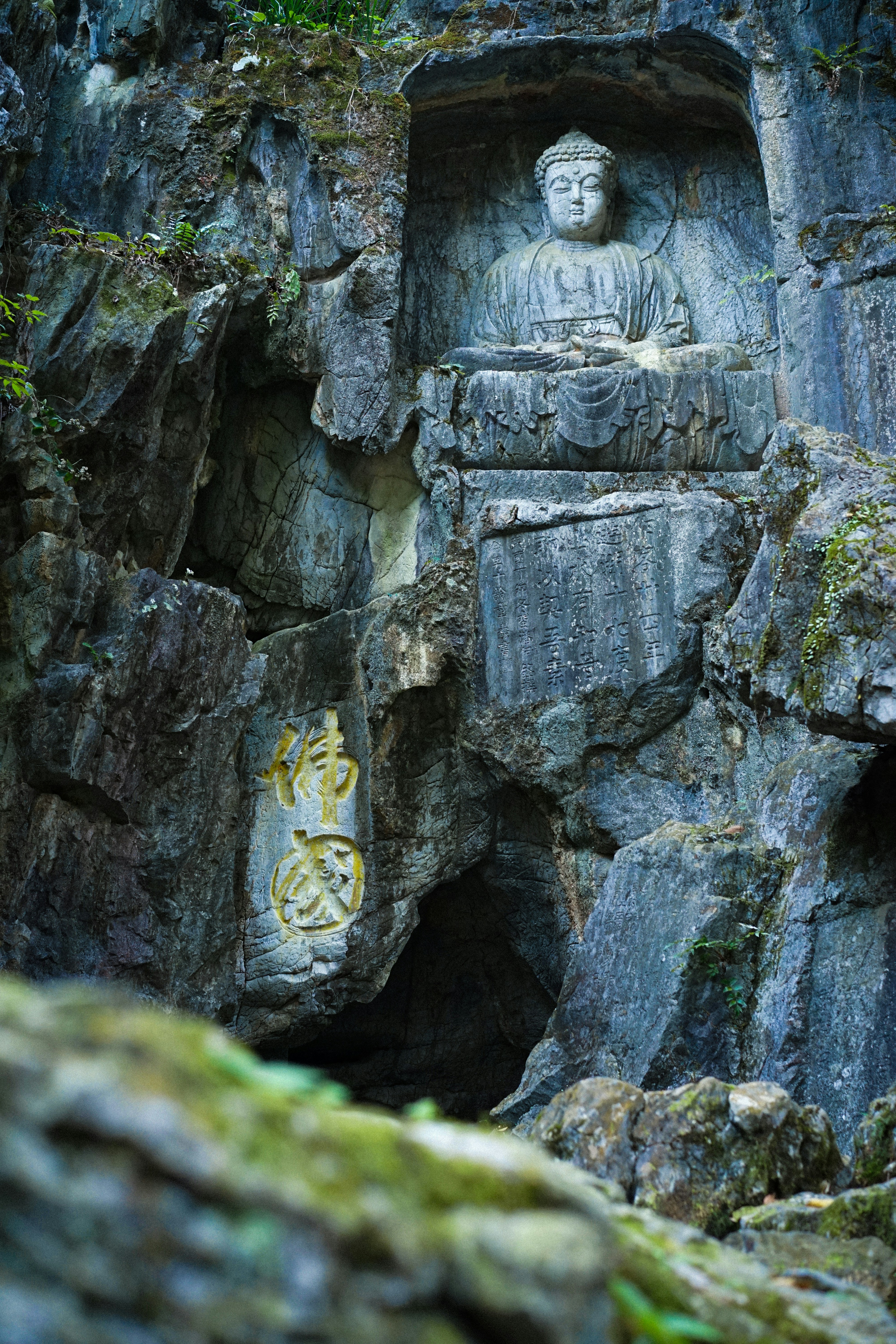 Buddha statue carved into rocky cliffside, surrounded by natural textures and inscriptions. The serene figure embodies a sense of tranquility amidst rugged terrain.