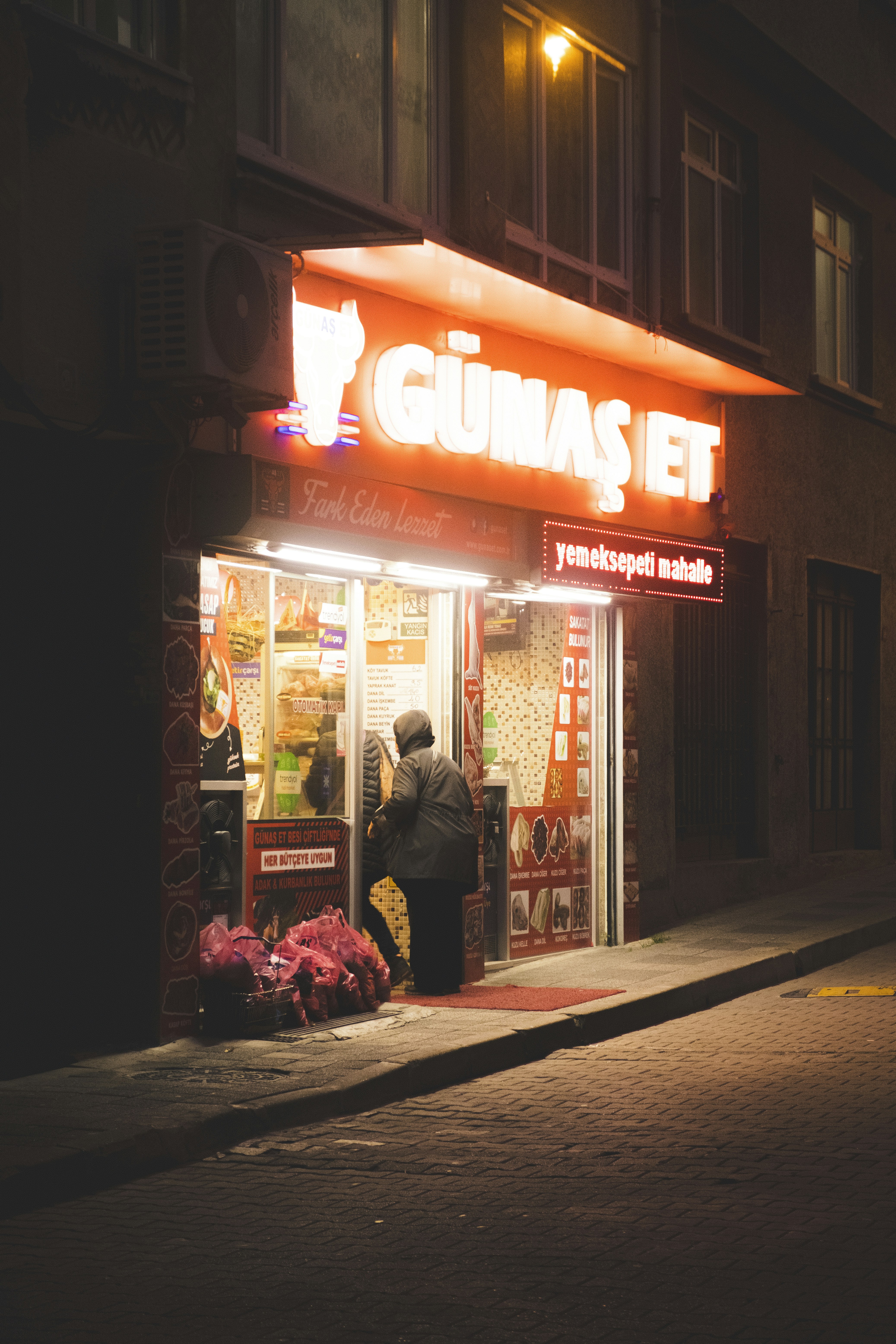 A warmly lit butcher shop on a dimly lit street, with a person entering under the neon sign.