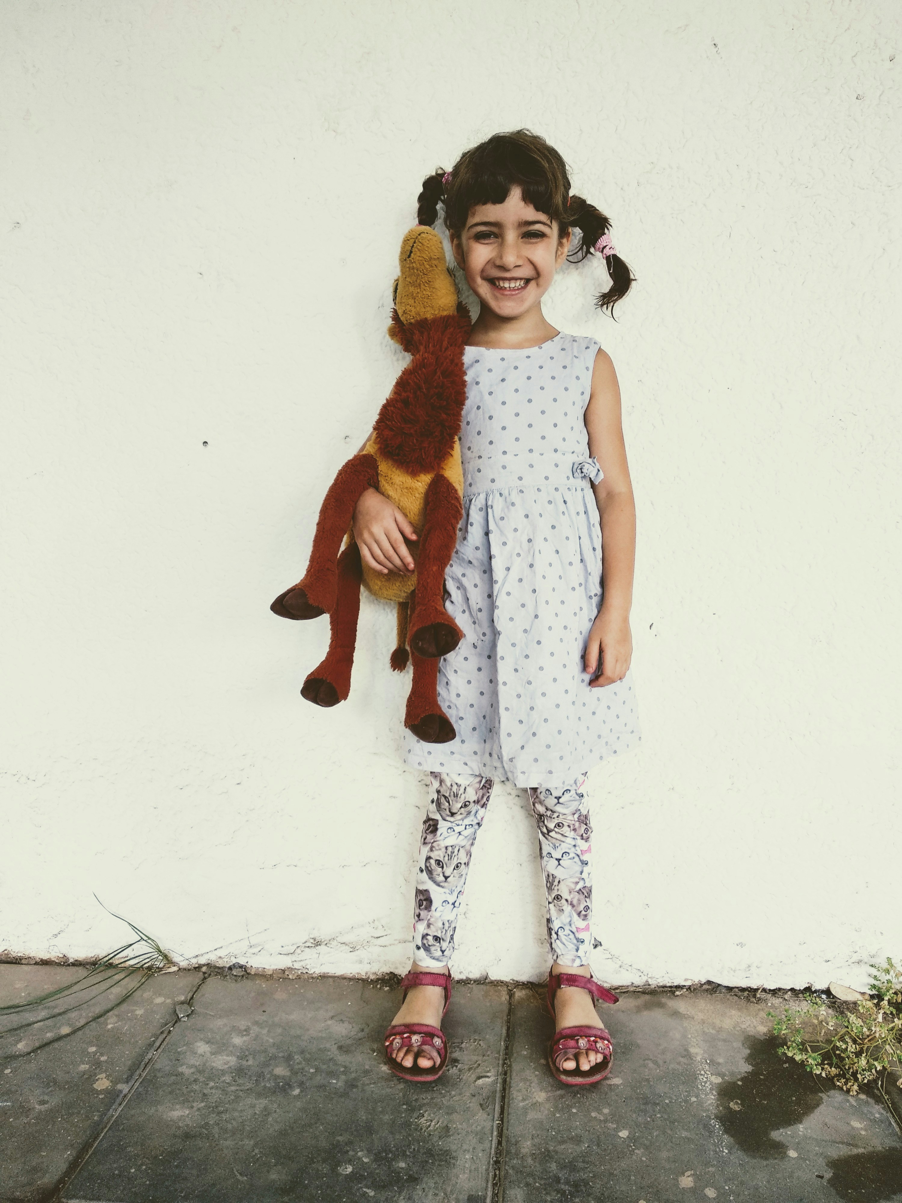Young girl in a dress and leggings smiles widely while holding a stuffed camel against a plain backdrop.