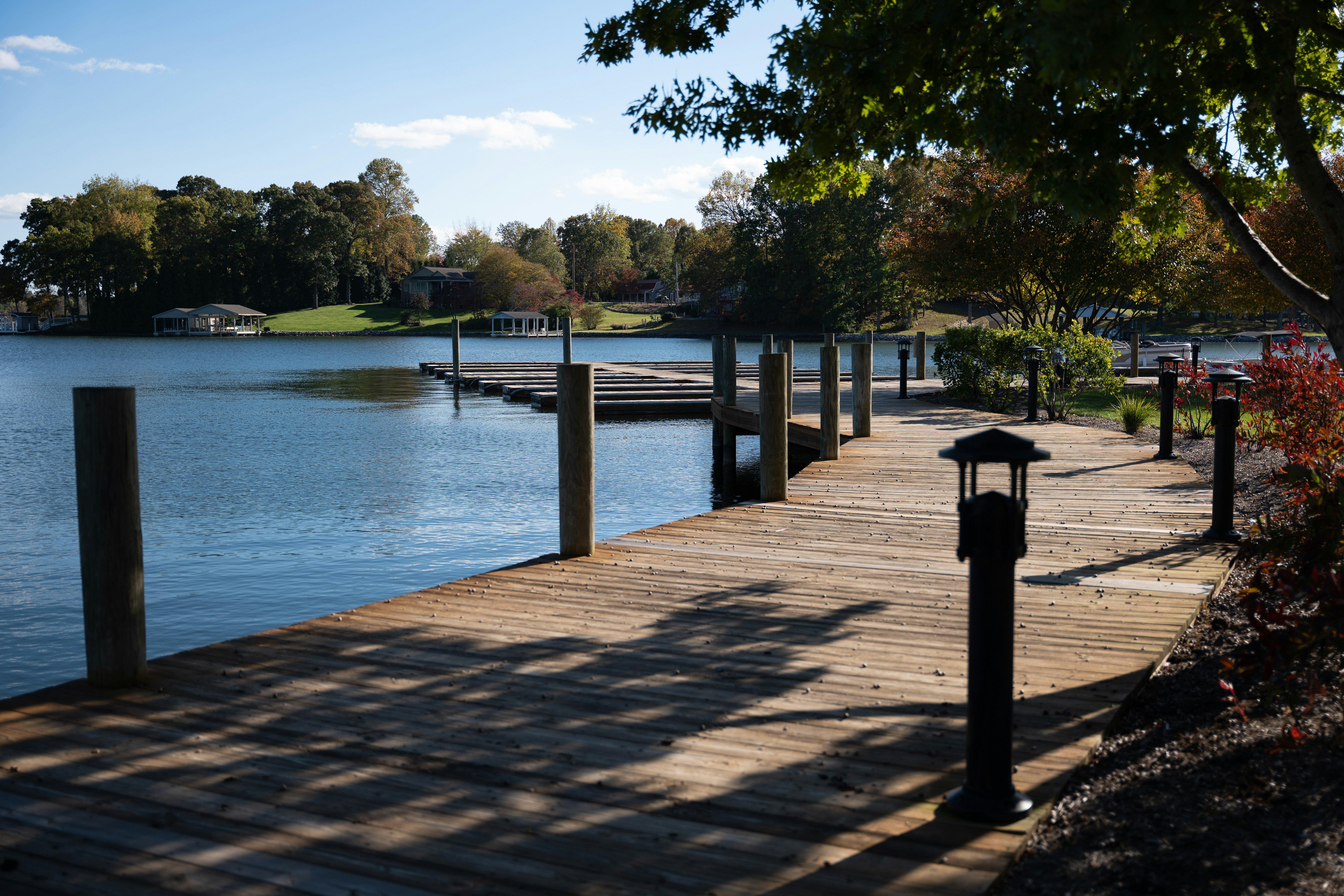 Sunlit boardwalk by a calm lake with shadows and autumn foliage