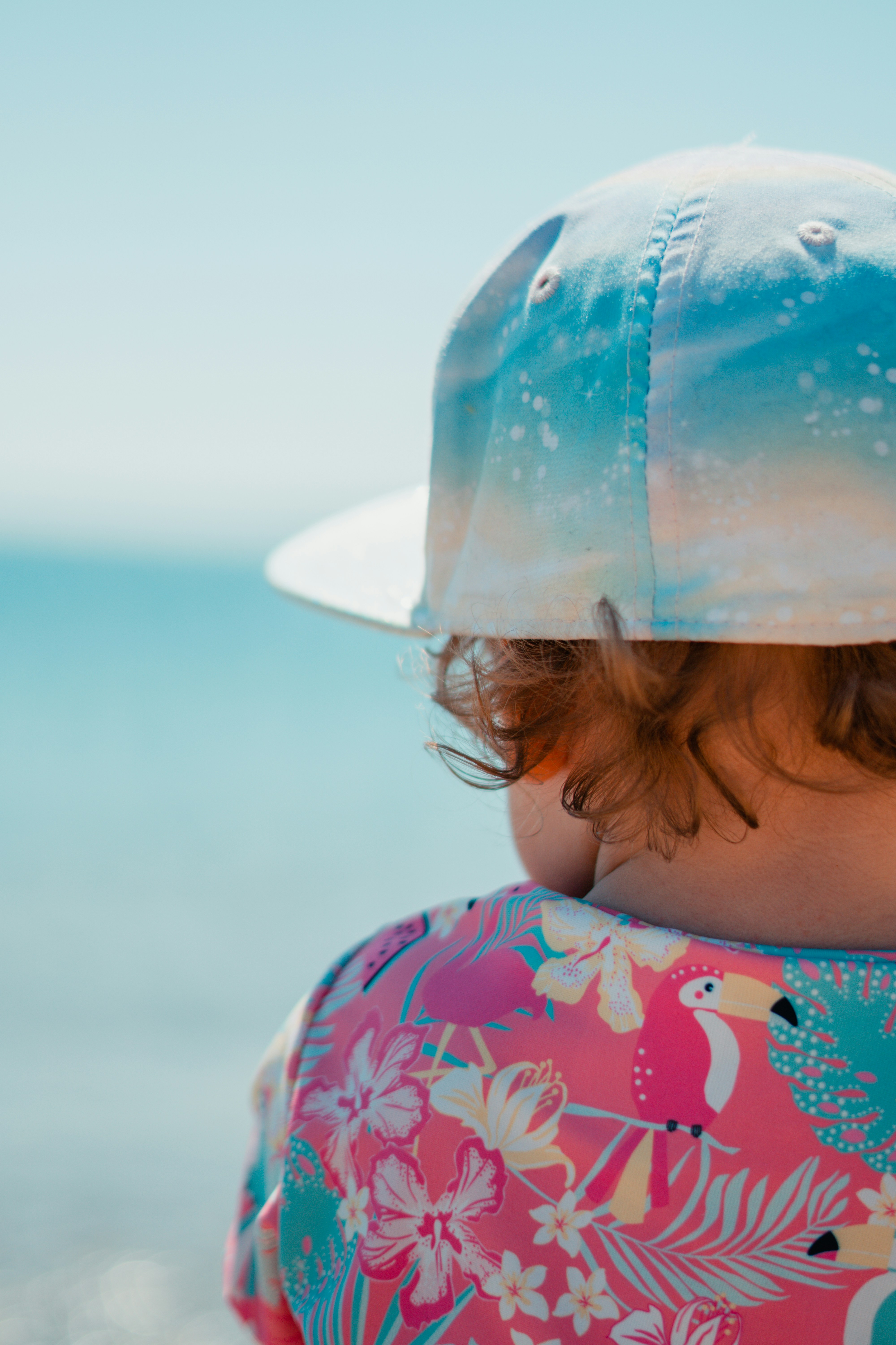 Child wearing a colorful floral shirt and a sun hat gazes at the tranquil ocean. The scene captures the essence of carefree summer days.