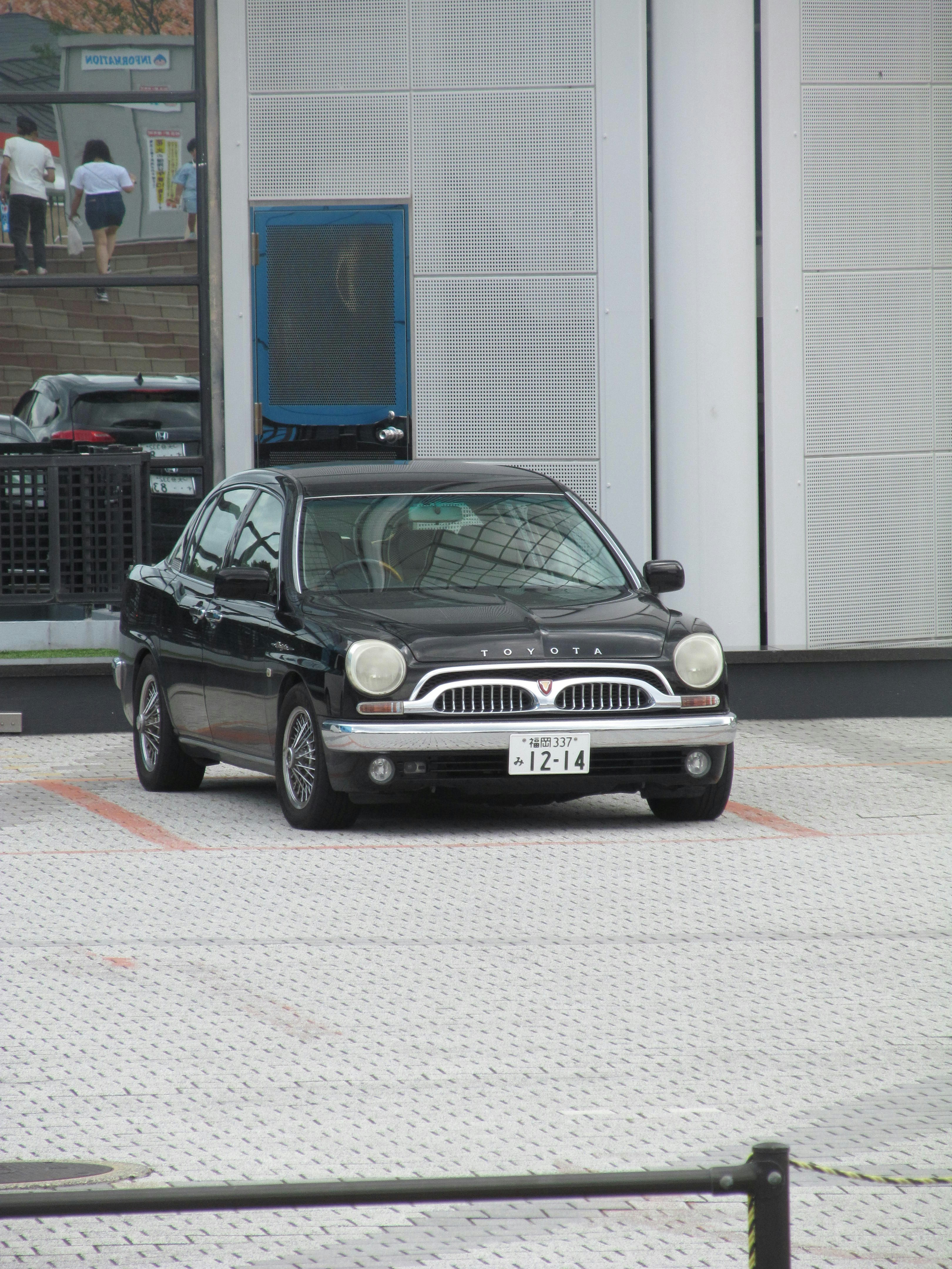 Photograph of a black Toyota sedan parked in a modern plaza with white panel walls and glass reflections. The chrome grille and license plate are clearly visible.