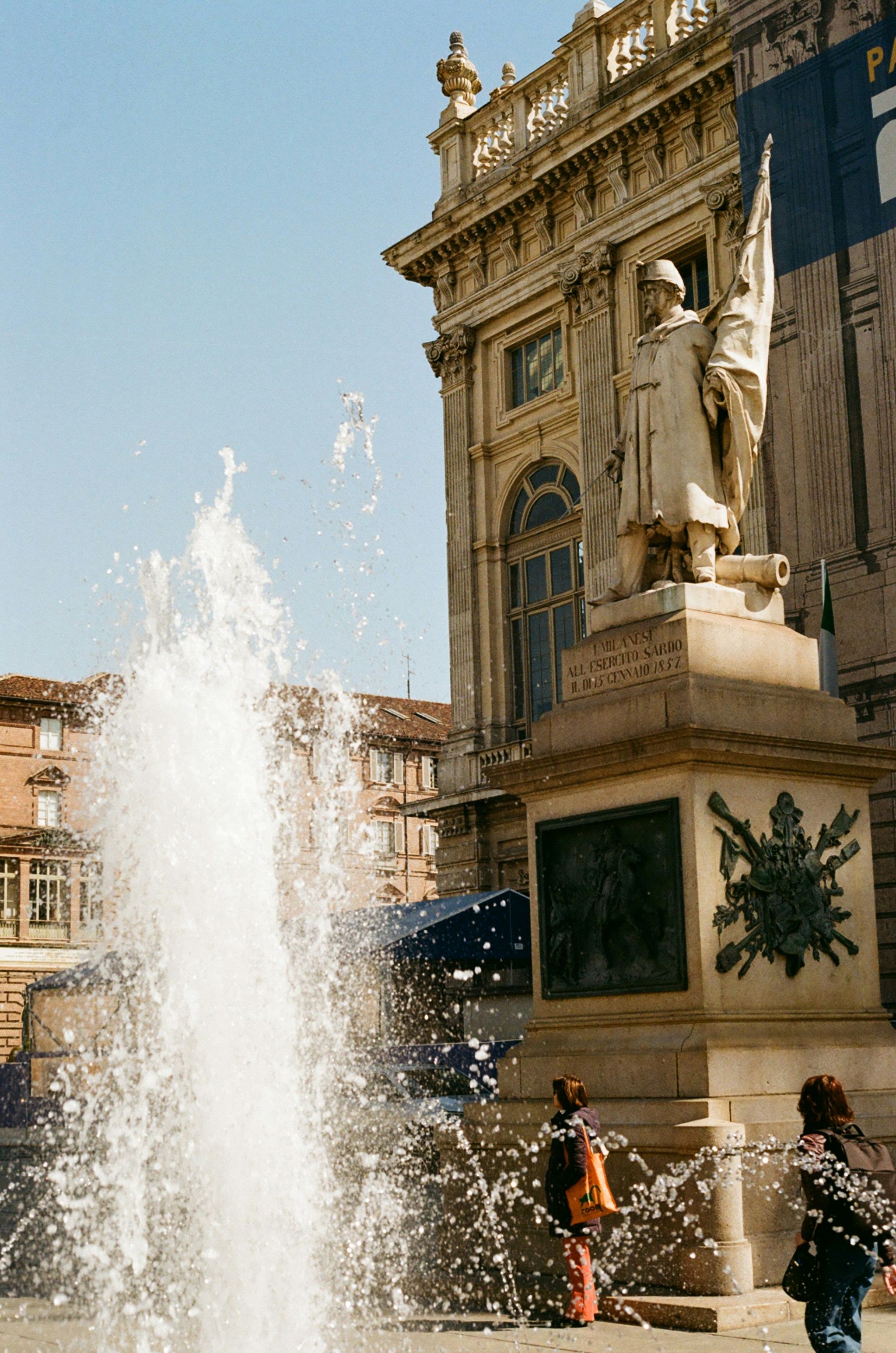 Sunlit plaza featuring a grand statue on a pedestal beside a central fountain, with pedestrians nearby.