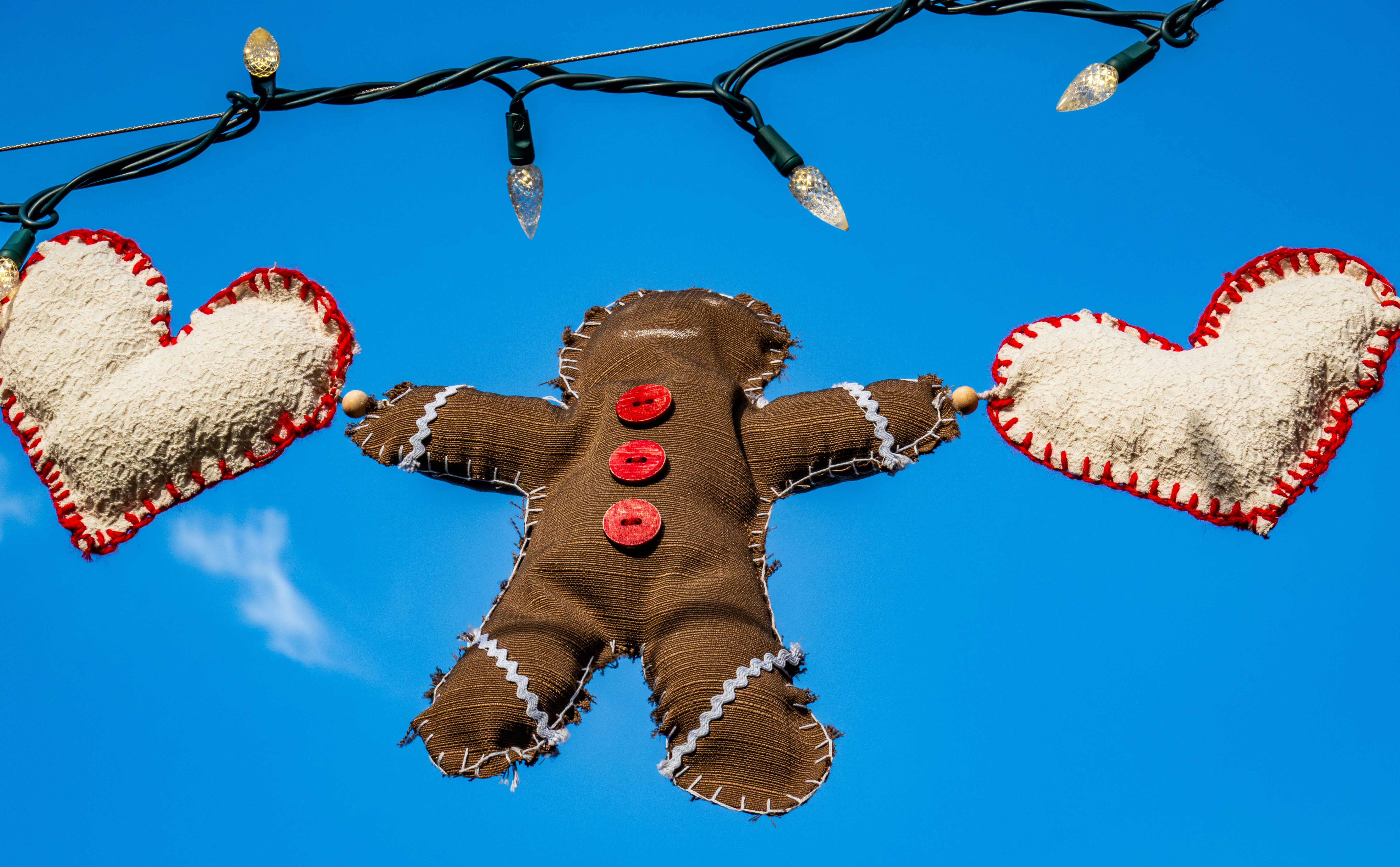 A festive gingerbread figure adorned with red buttons, flanked by two stitched hearts, suspended against a clear blue sky.