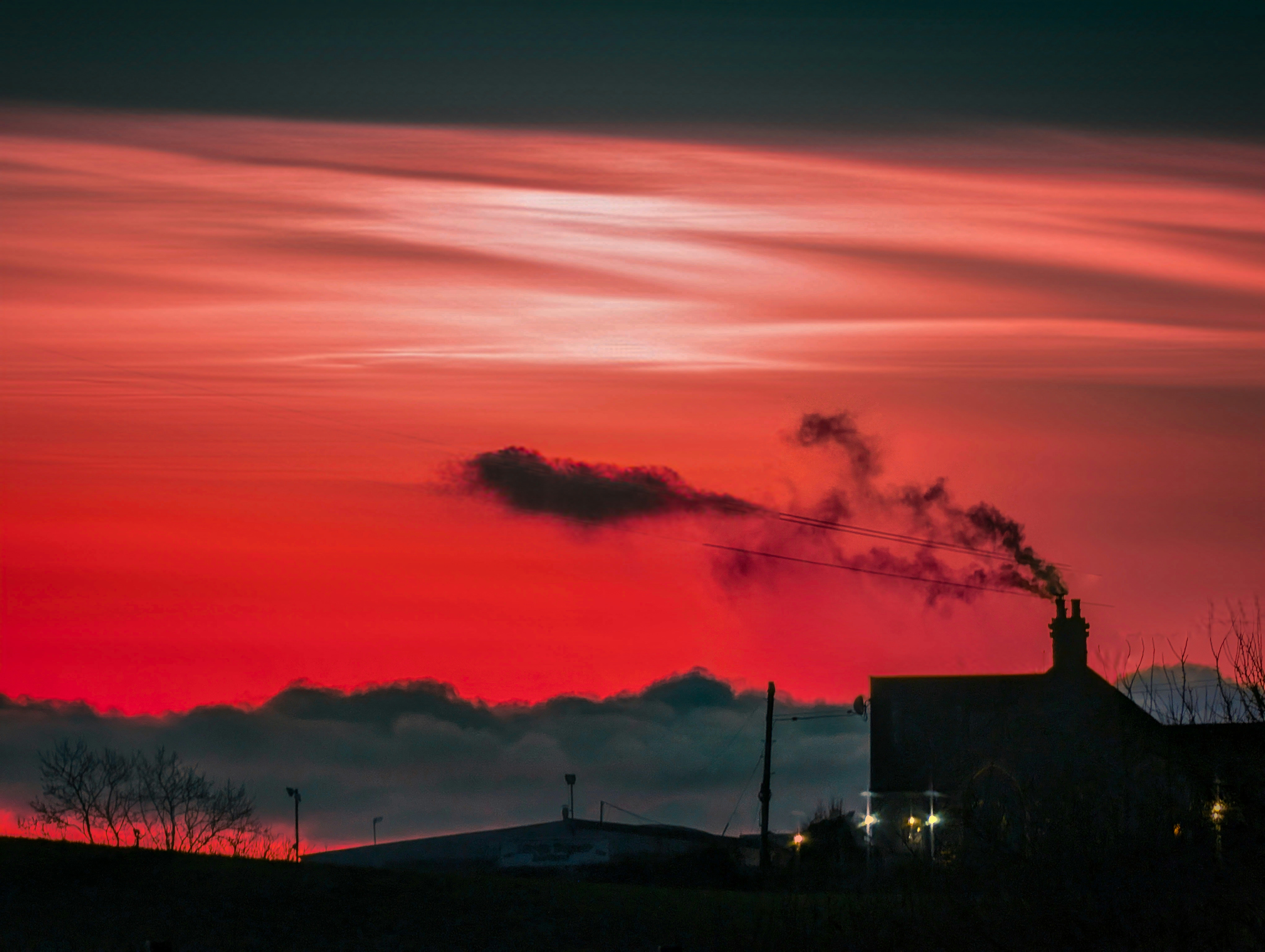 A silhouette of a farmhouse with a smoking chimney stands against a vivid red sunset and layered clouds. This landscape photograph captures a quiet rural horizon at dusk.