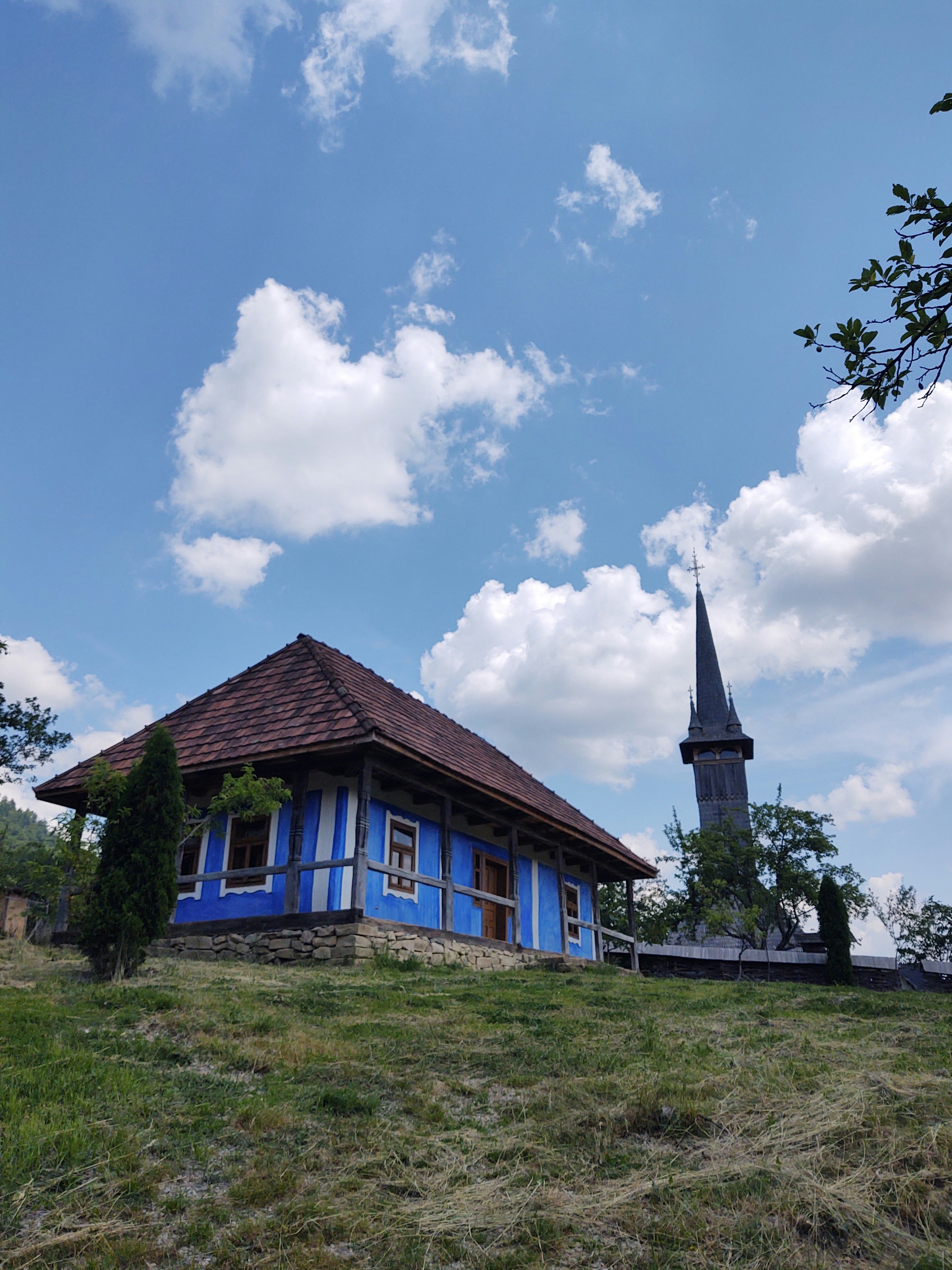 Traditional blue and wooden house with a tall spire in the background, surrounded by greenery and a bright sky with fluffy clouds.