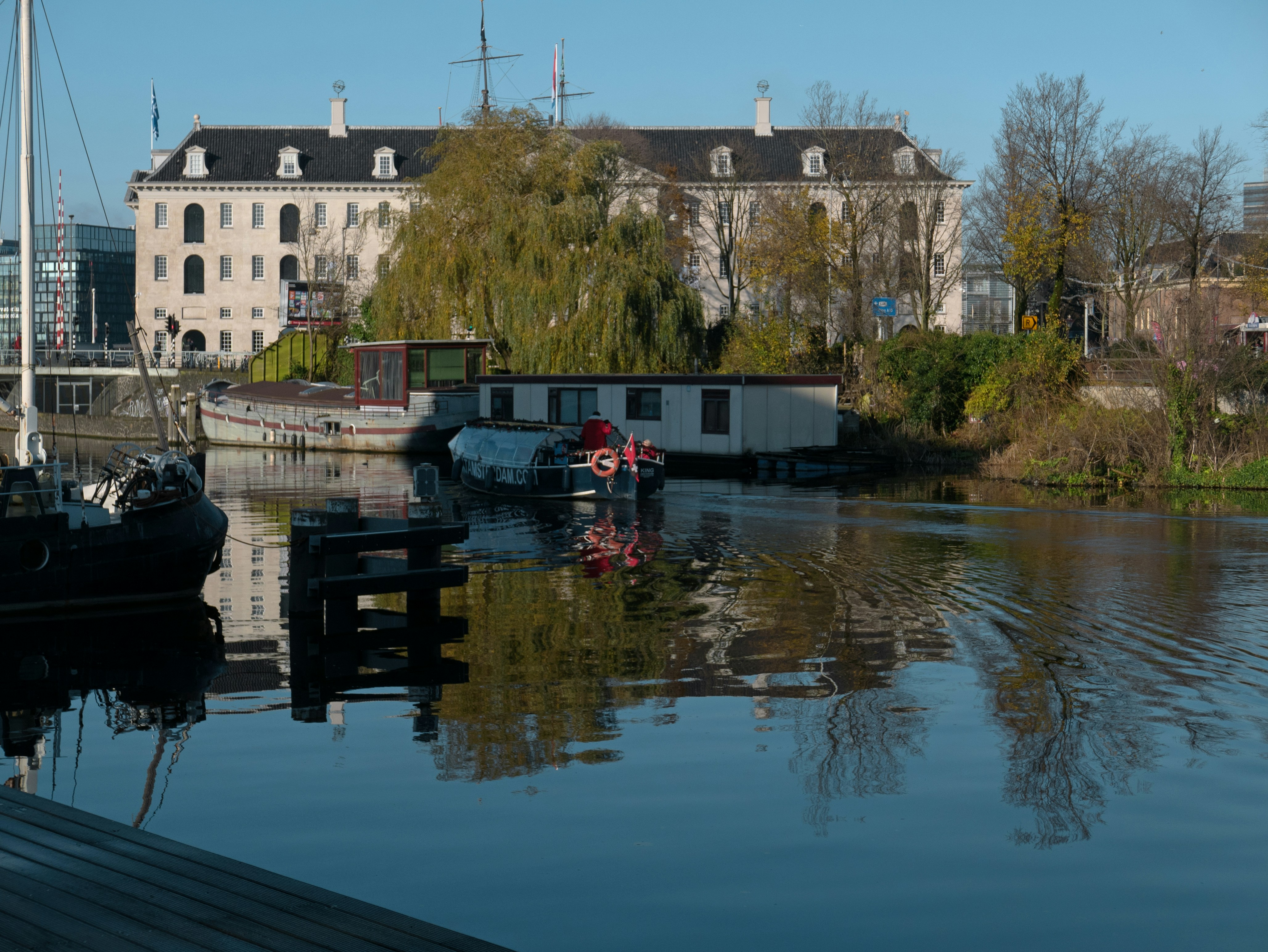 Free photo of reflecting water in the canal Nieuwe Vaart under a blue sky. In the background the historical building facade of the contemporary Maritime Museum in Amsterdam city. Some houseboats are drifting in the canal water. Street photography of cities in The Netherlands by Fons Heijnsbroek in free 2023 // Gratis foto Amsterdam, met uitzicht over het water van de Nieuwe Vaart op woonboten en op de oude stenen gevel van het historische Scheepvaartmuseum. Herfst met veel zon en weinig wind; dus veel weerspiegelingen en schaduwen in het gladde water; Amsterdam stad, fotografie Nederland.