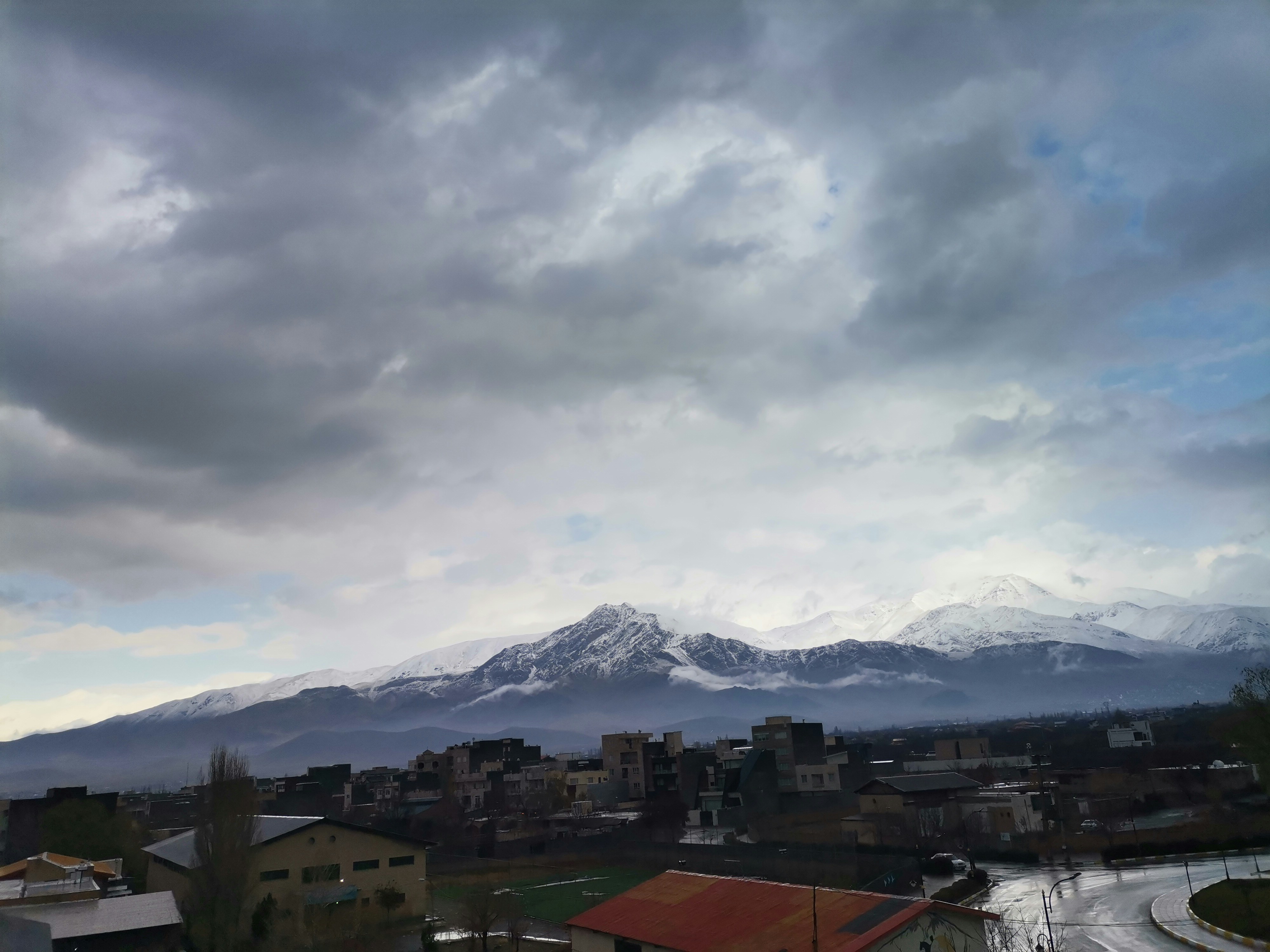 Snow-capped mountains rise behind a low city skyline as moody clouds drift overhead.