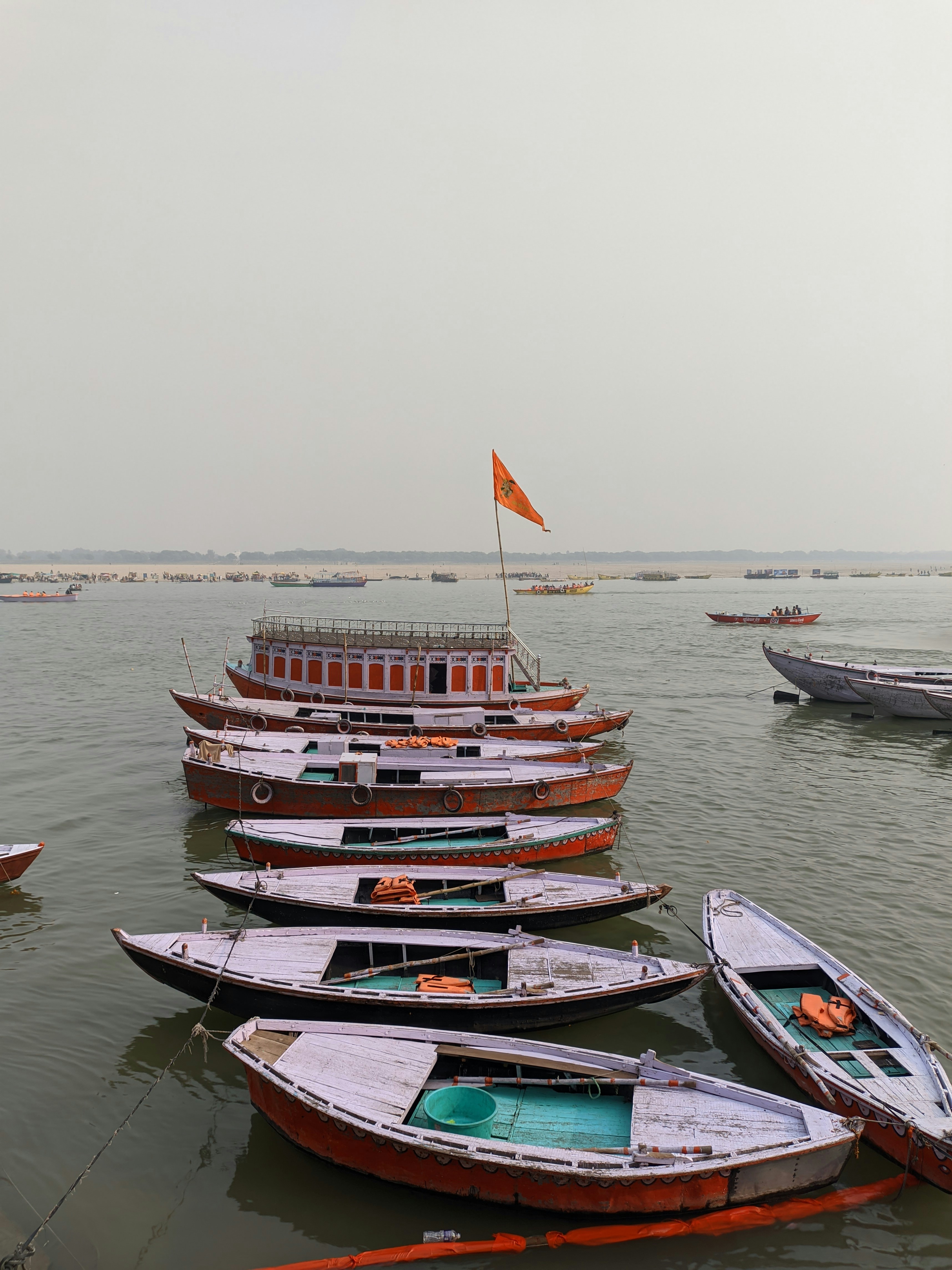 Colorful boats docked along a calm river with an orange-flagged vessel leading the line.
