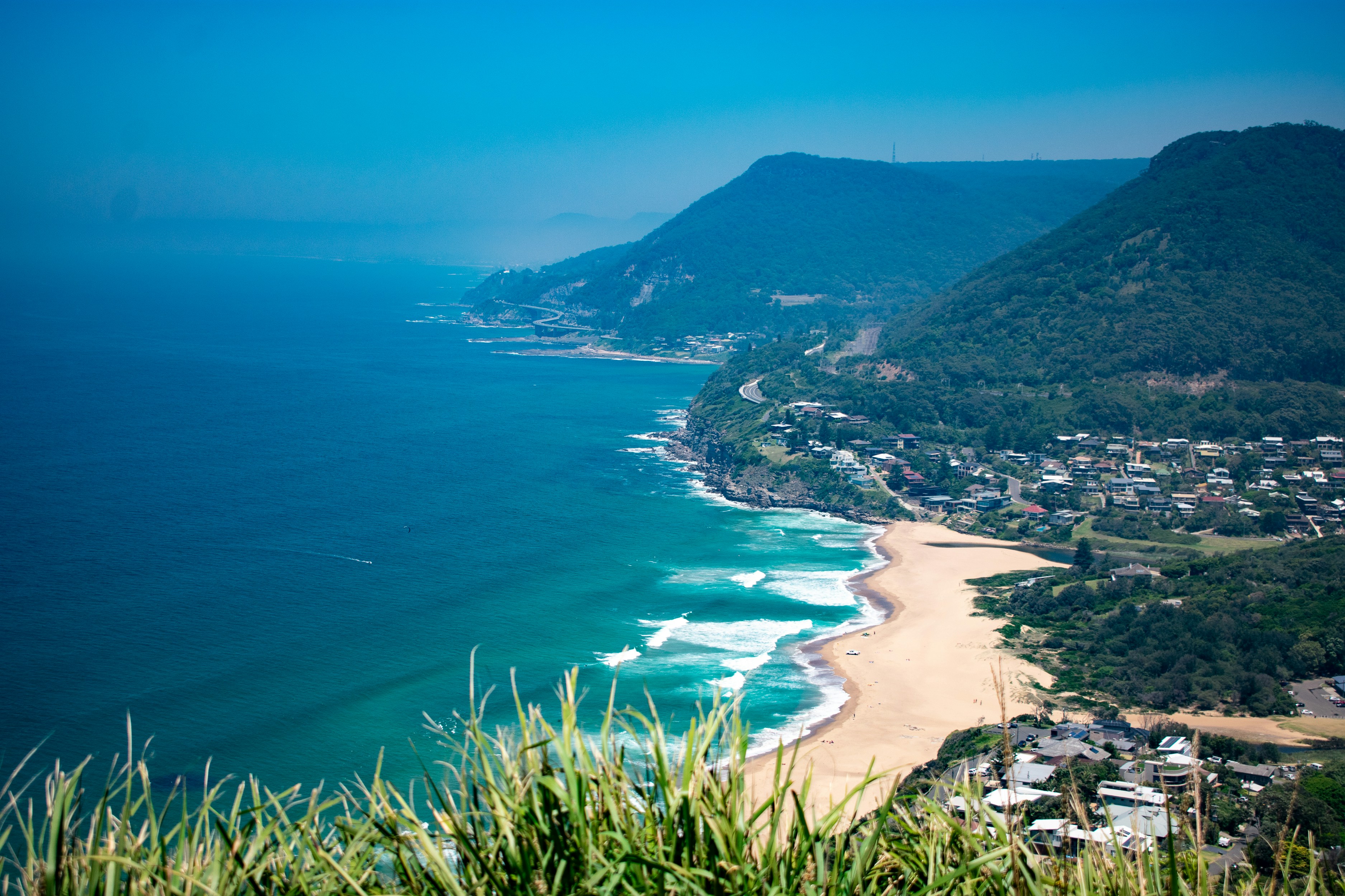 Sandy beach and turquoise waves viewed from a grassy hill with distant mountains under a clear blue sky.