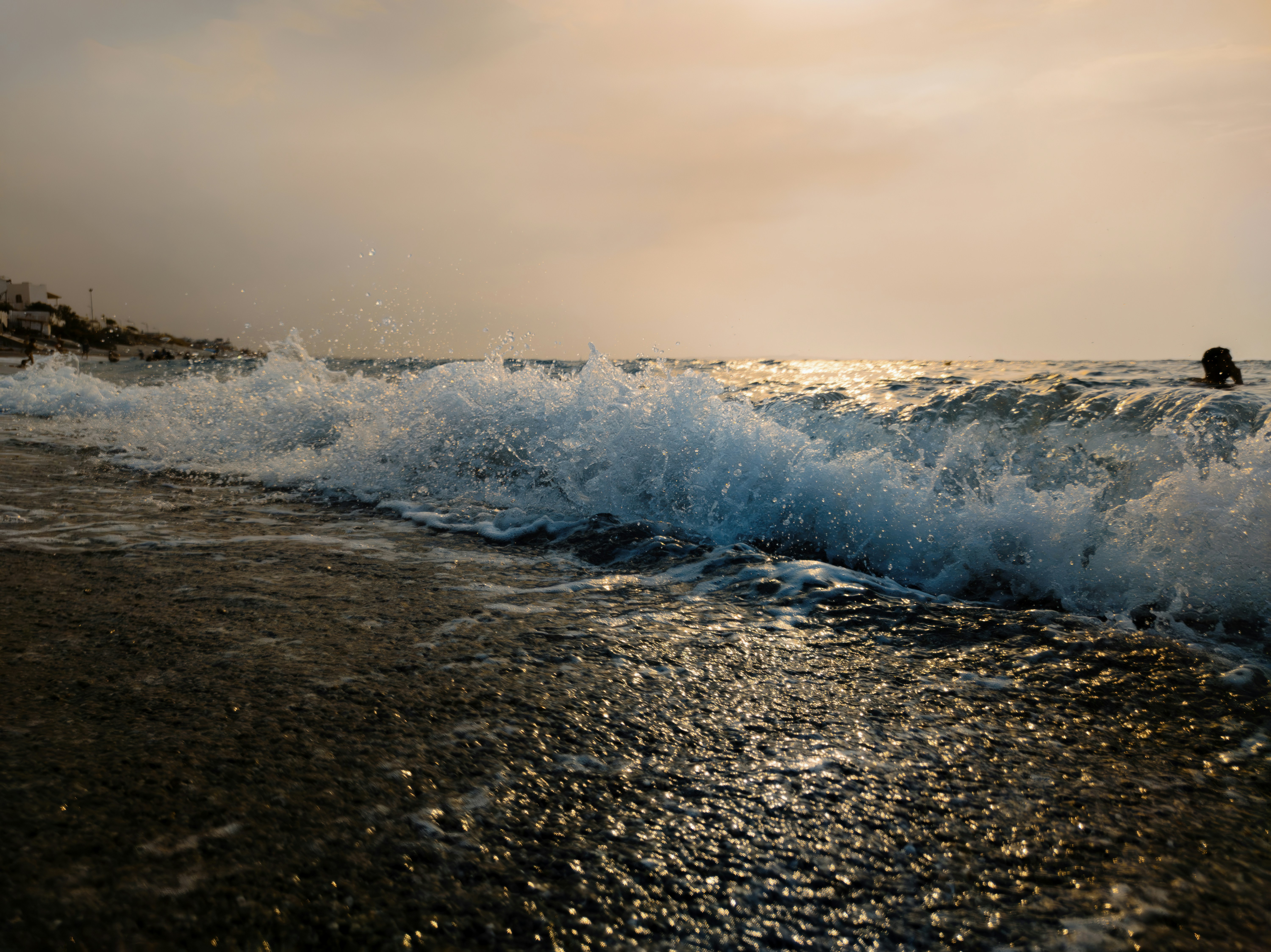 Golden-hour photograph of breaking waves along a rocky shoreline, with a distant silhouette against the horizon. The scene emphasizes motion and warm reflections on the water.