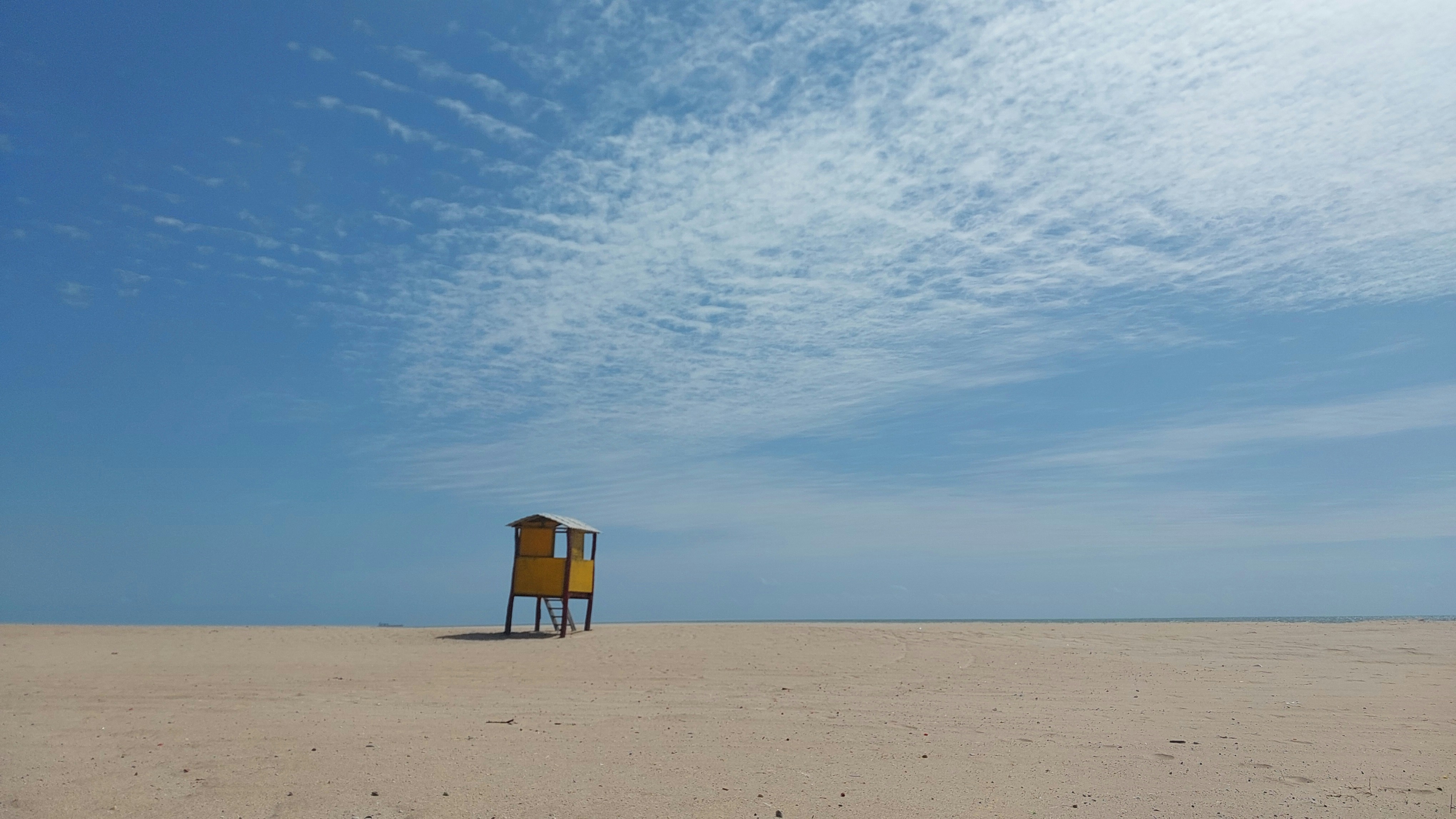 Yellow lifeguard chair stands alone on a vast sandy beach under a wide blue sky.
