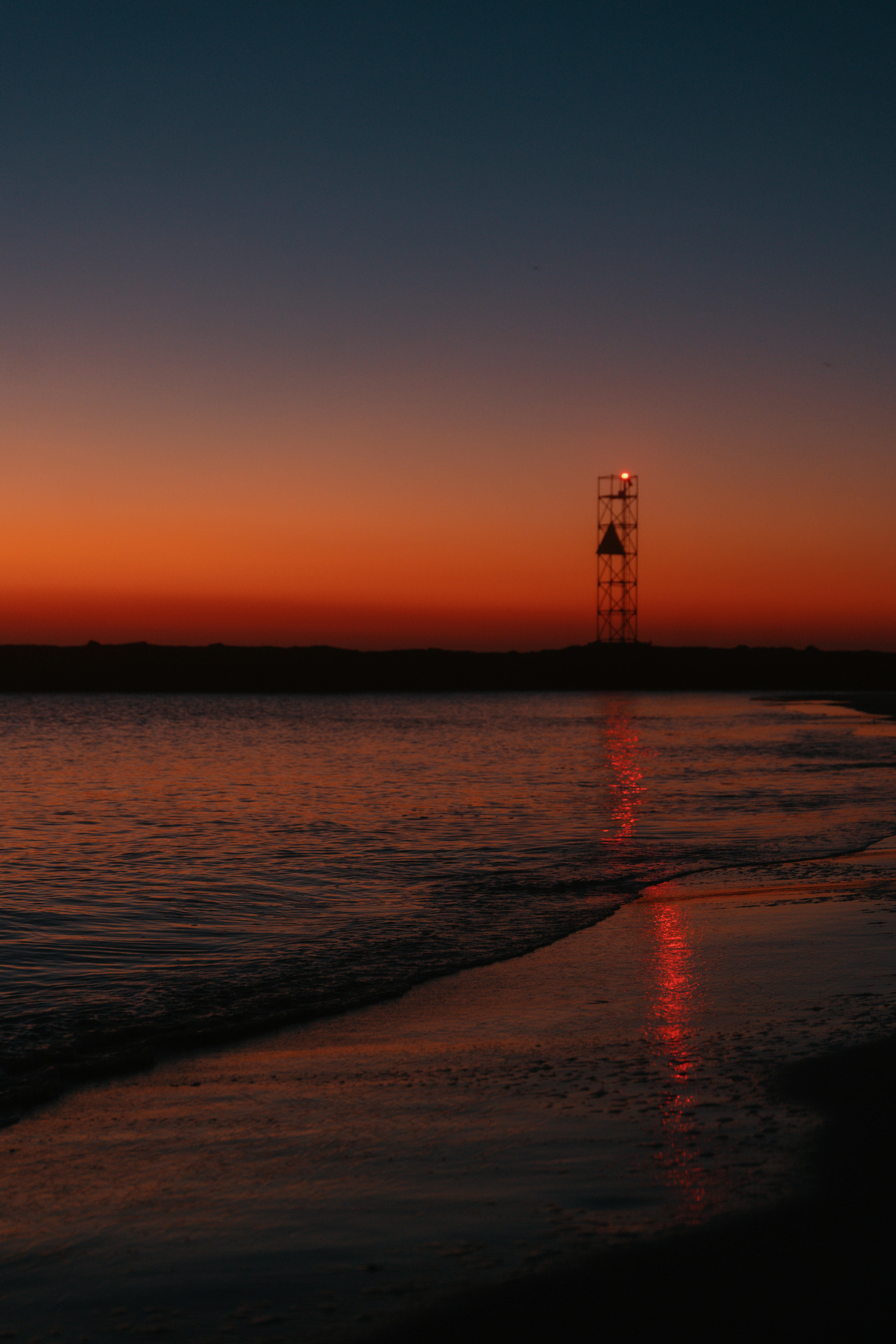 Silhouetted lighthouse with its reflection stretching across the calm water at sunset.