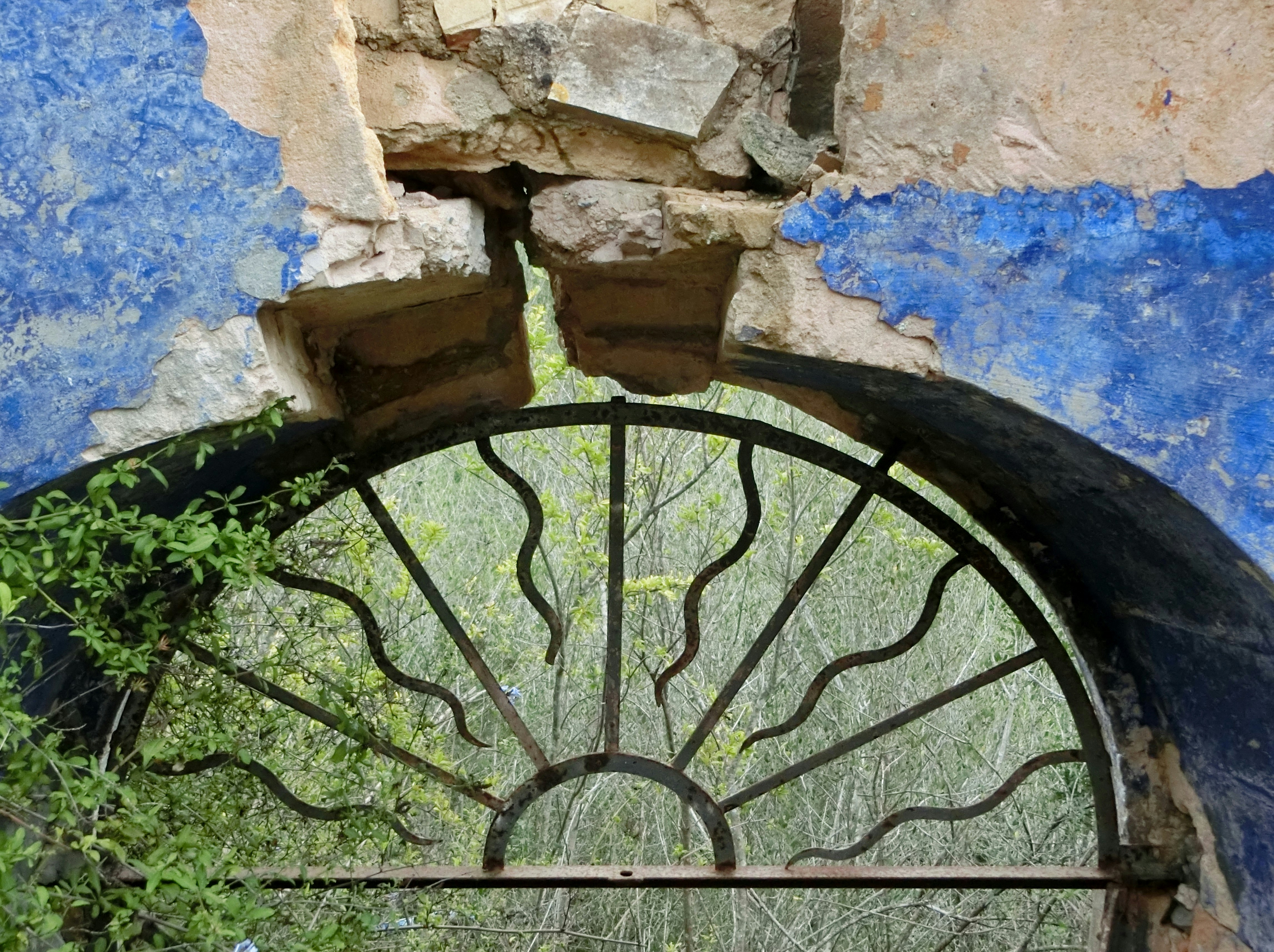 Photograph of a circular wrought-iron sunburst grille behind a crumbling blue wall arch, with greenery at the edges.