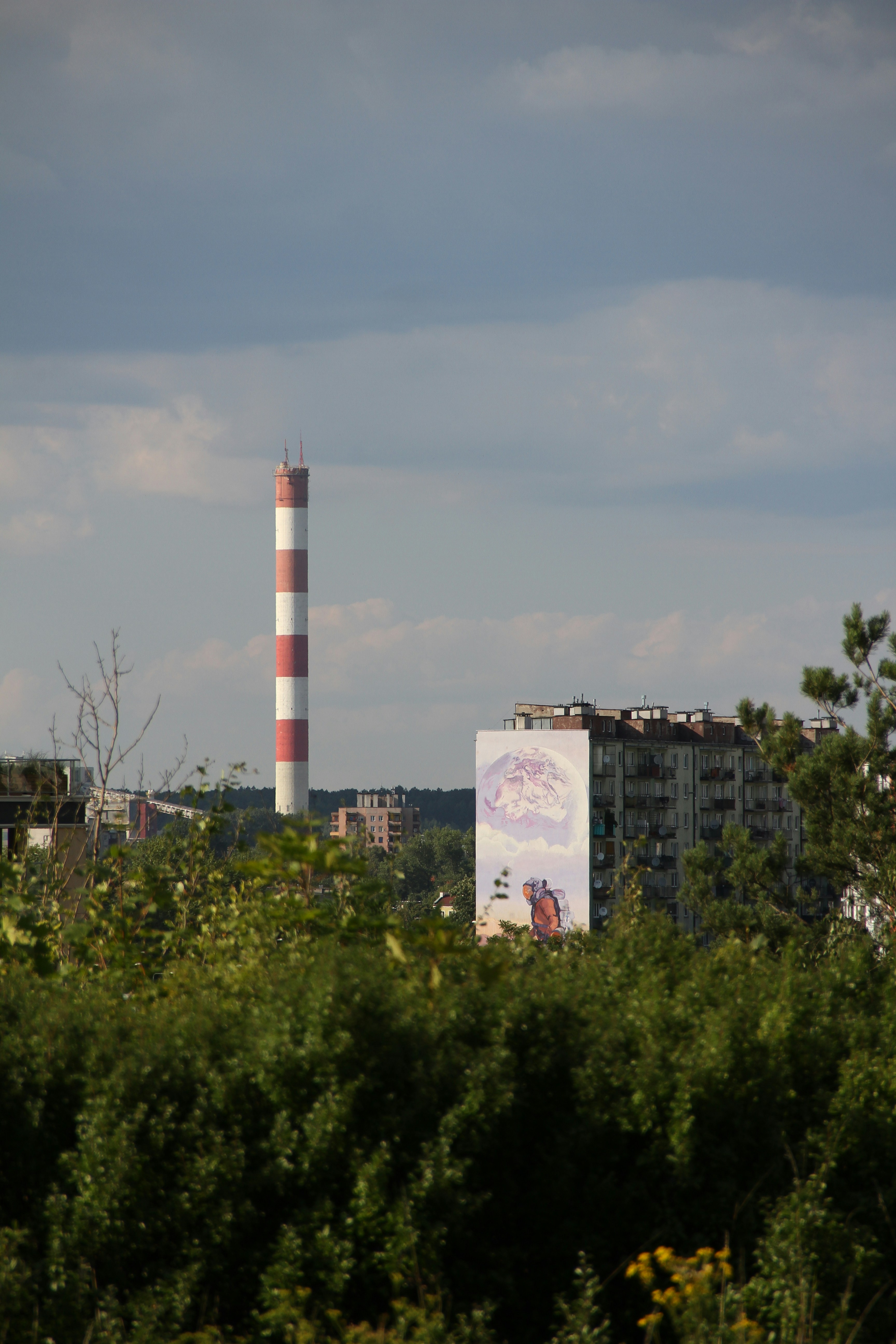 Industrial chimney rises above a green fringe, with a mural-faced building in the mid-ground