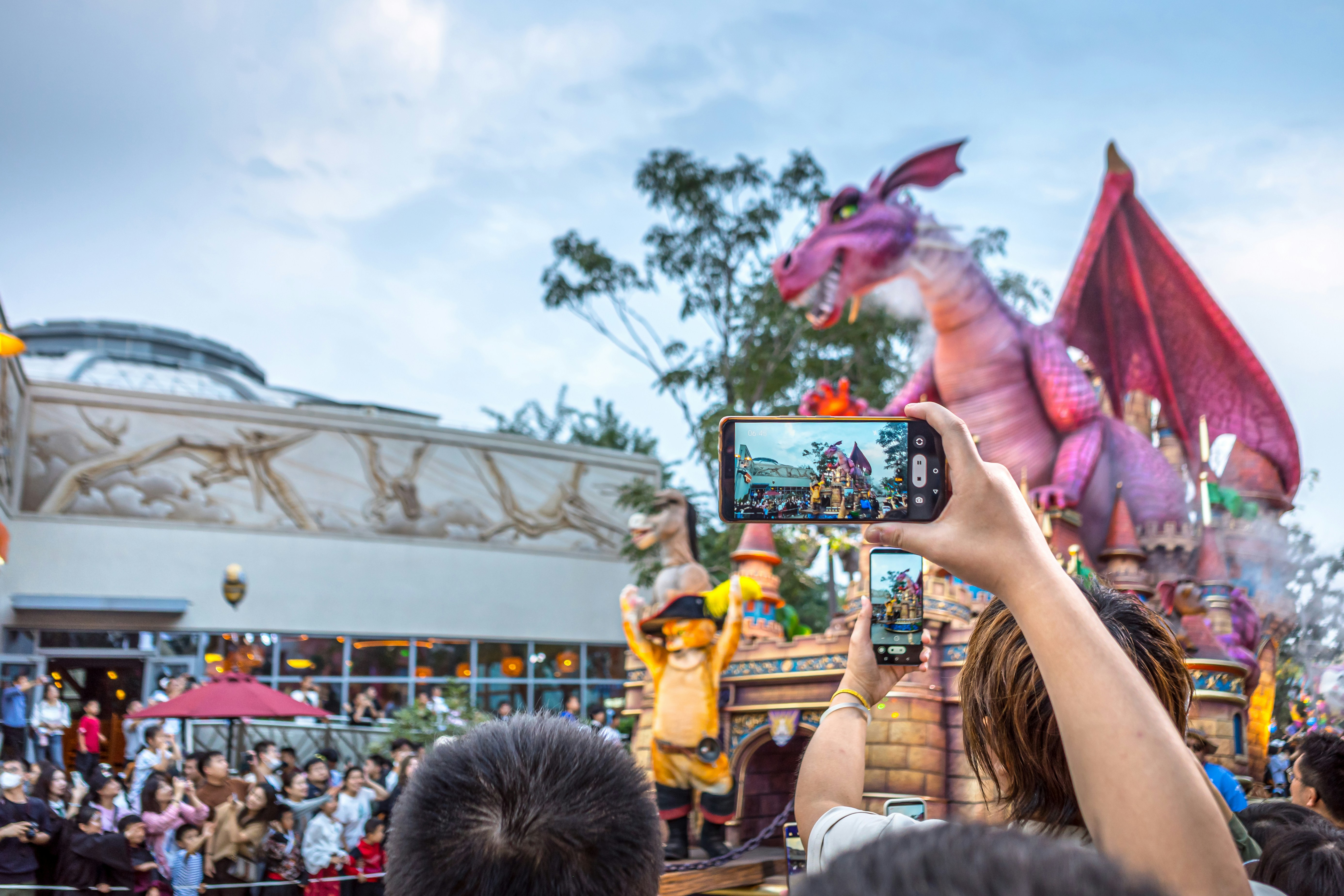 Crowd-packed street festival with a vibrant pink dragon float dominating the scene. In the foreground, a person holds up a smartphone to capture the moment.