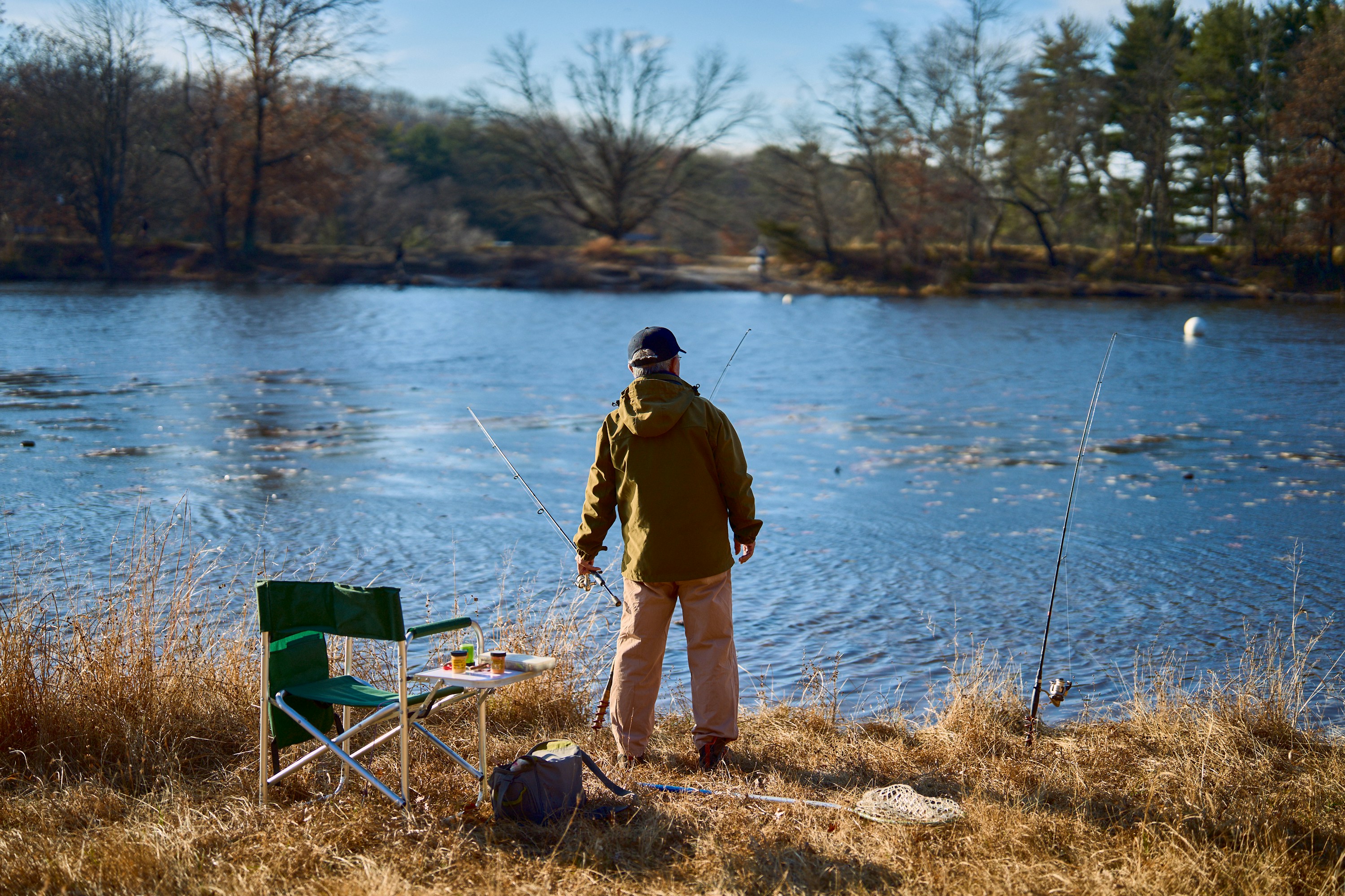 Person in a brown jacket stands beside a lake with a folding chair and tackle box on a sunny day.