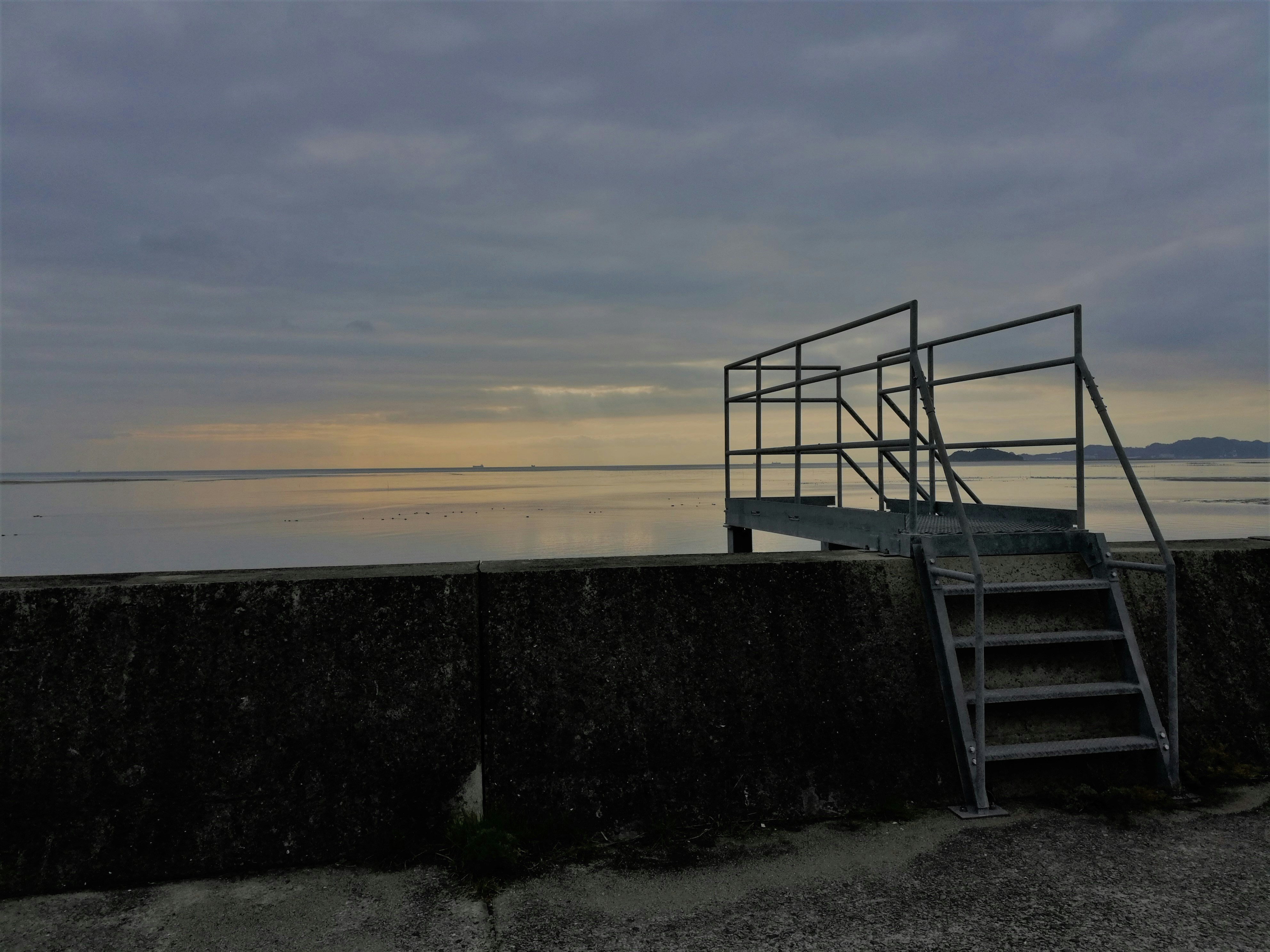 Metal stairs leading over a concrete wall to calm waters under a cloudy sky.