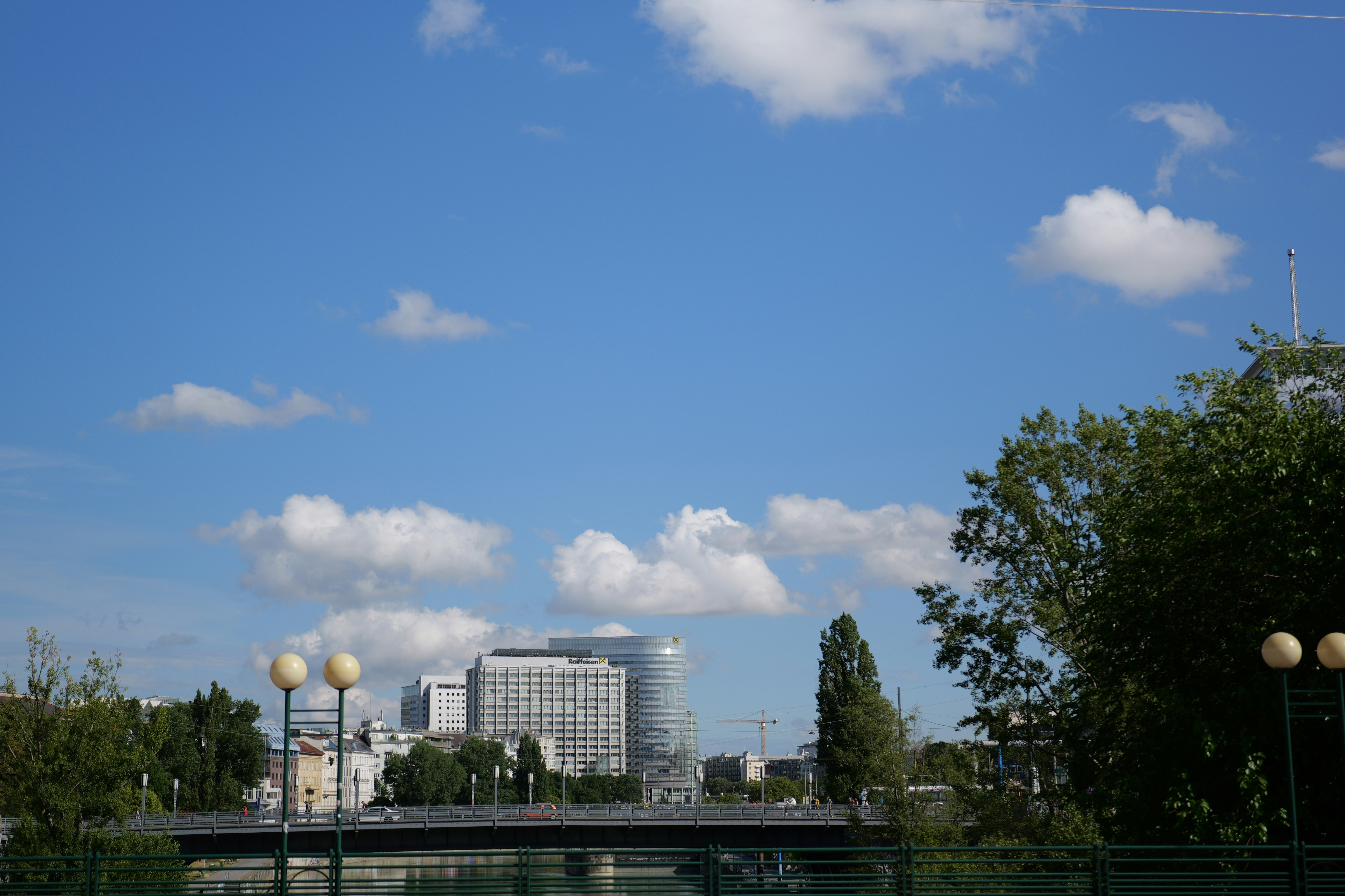Cityscape with modern buildings across a river, framed by lush green trees under a vibrant blue sky with scattered clouds.