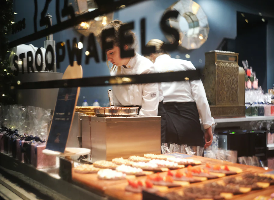 Two chefs work behind a glass display in a bakery or dessert shop, with trays of pastries in the foreground. Reflections and signage on the glass add a busy indoor street-side feel.