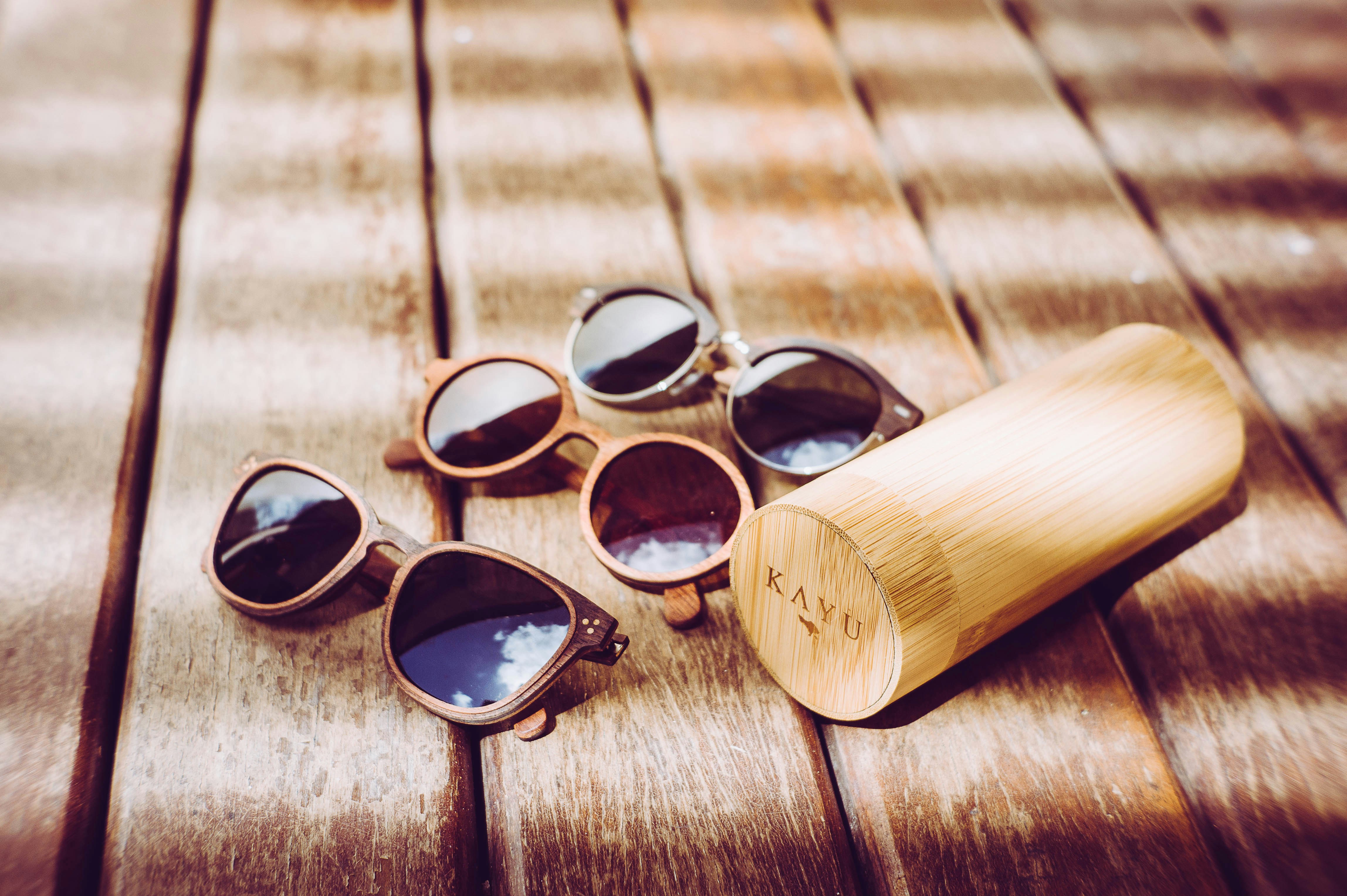 Several sunglasses scattered on a wooden deck beside a light wood capsule, casting soft shadows; a still-life study emphasizing texture and warm tones.