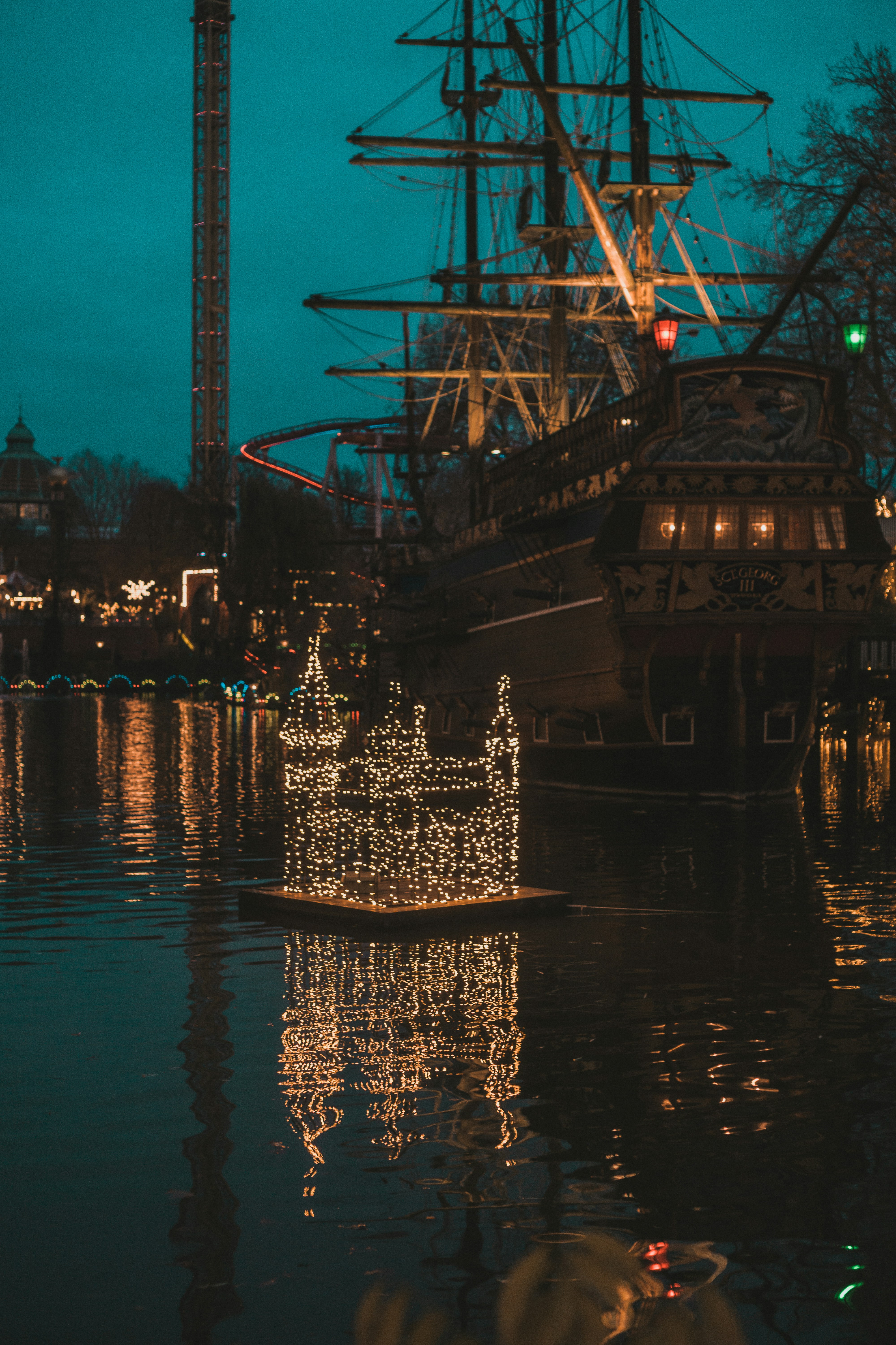 A beautifully illuminated structure resembling a castle floats on the water, reflecting the lights of a nearby ship and festive decorations around the harbor.